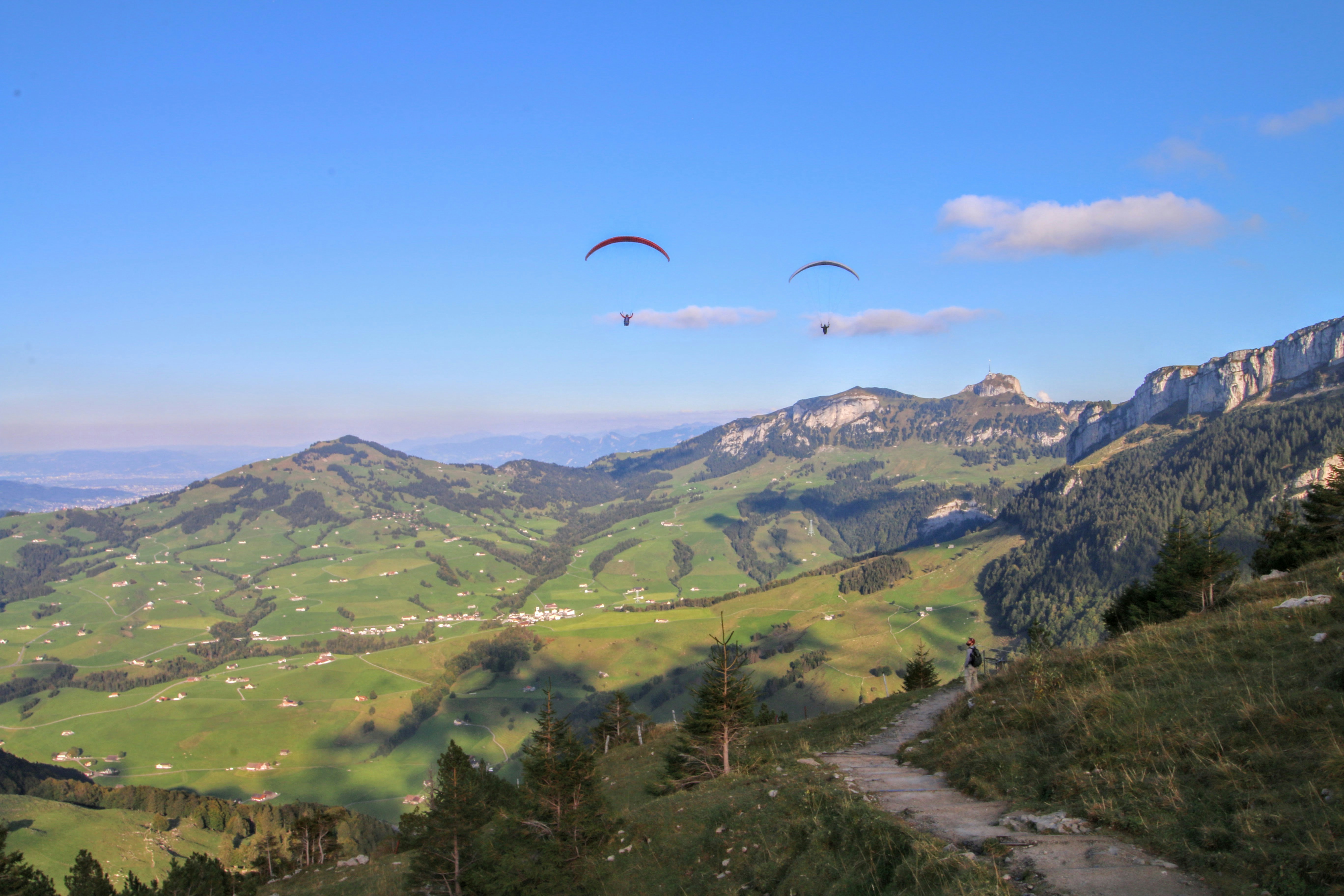 Two paragliders tracing arcs over a sunlit Alpine valley