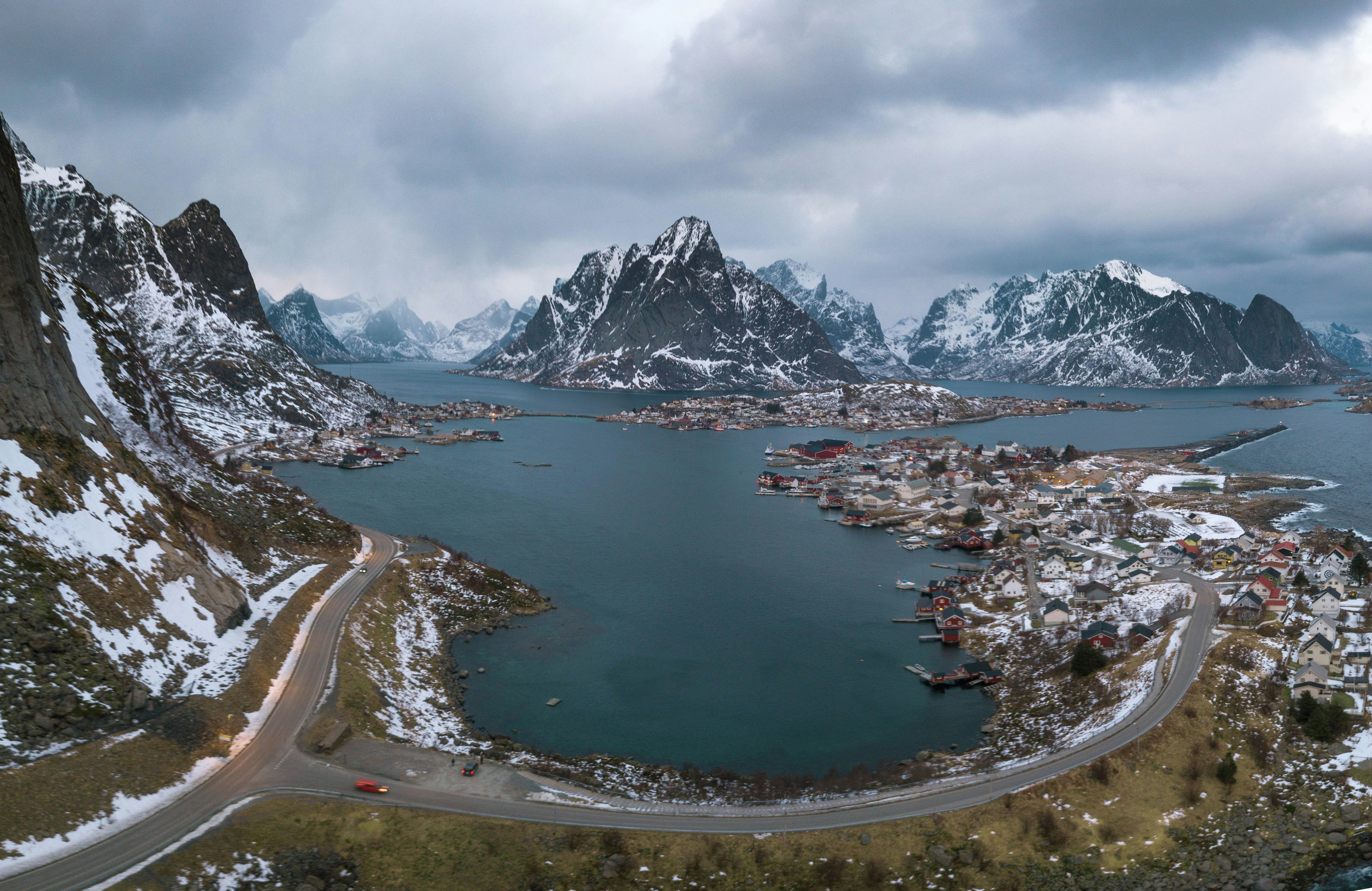Panoramic view of a snow-covered coastal town nestled between rugged mountains and a vast fjord under a cloudy sky.