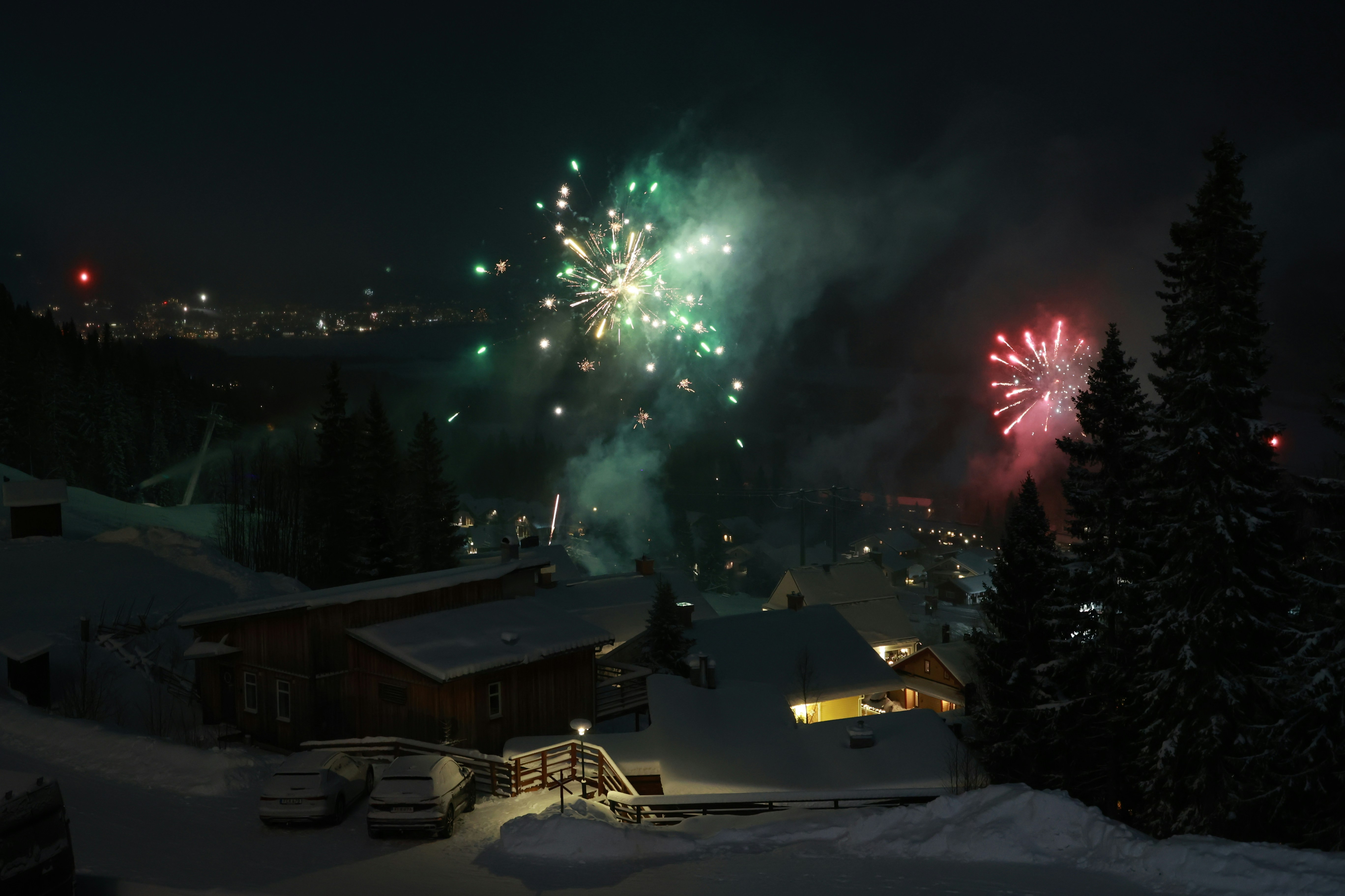 Colorful fireworks illuminate a snowy landscape at night, with cozy cabins nestled in the foreground. A festive atmosphere is created against a backdrop of dark skies.