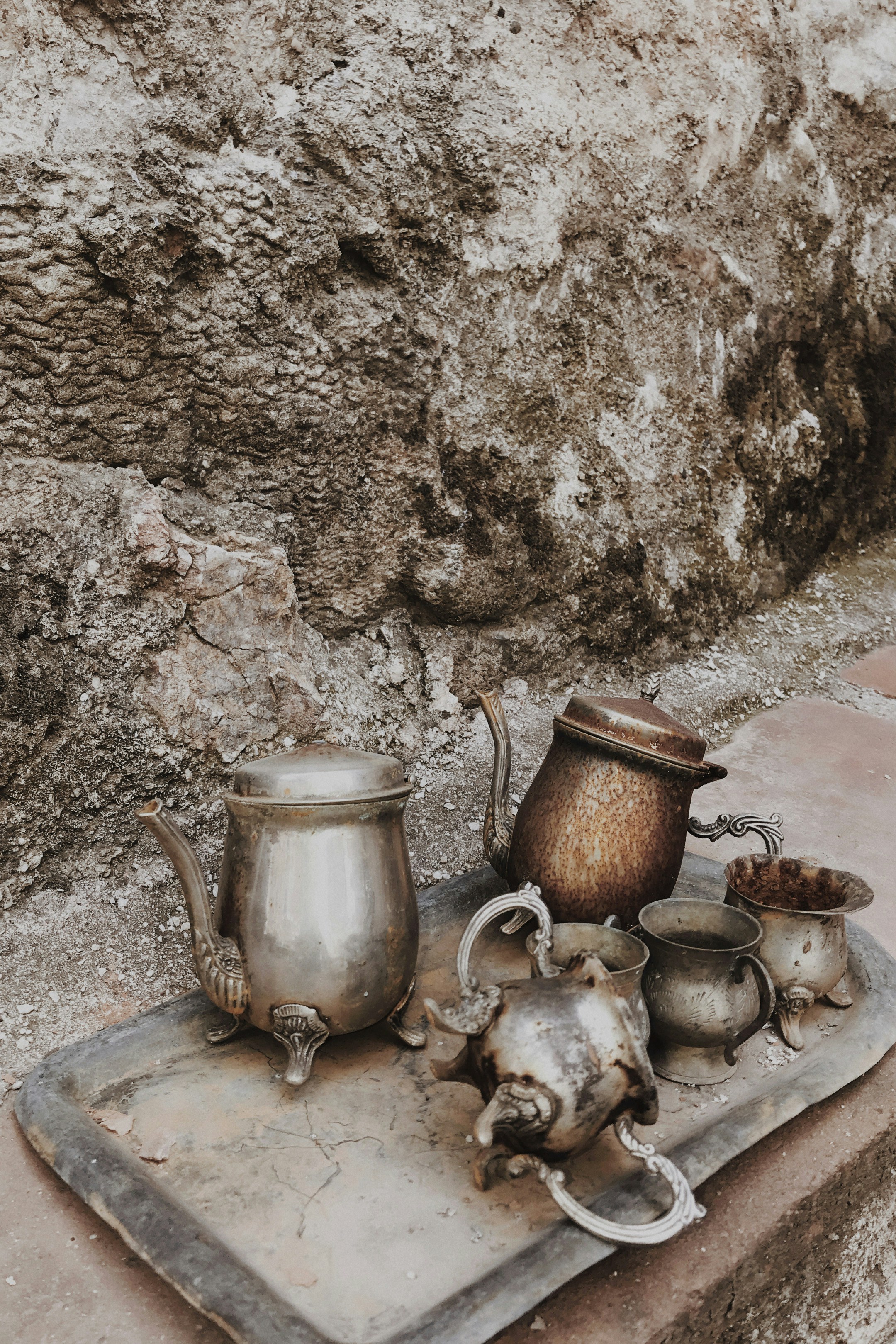 An antique silver tea set displayed on a weathered tray against a rugged stone backdrop. Each piece reflects a history of craftsmanship.