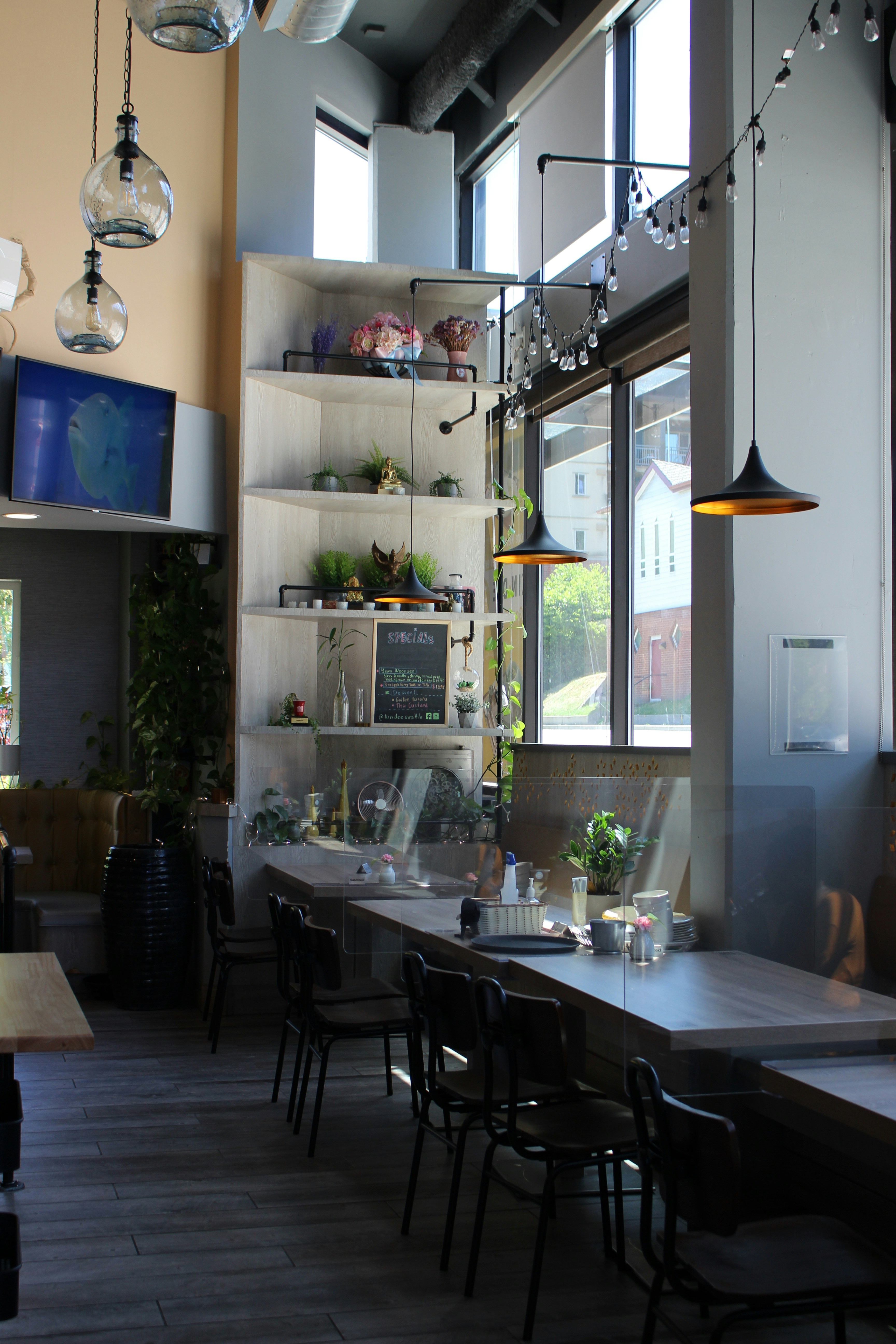 Modern café interior featuring a tall shelf adorned with plants and decorative items, alongside a long communal table and stylish pendant lights.
