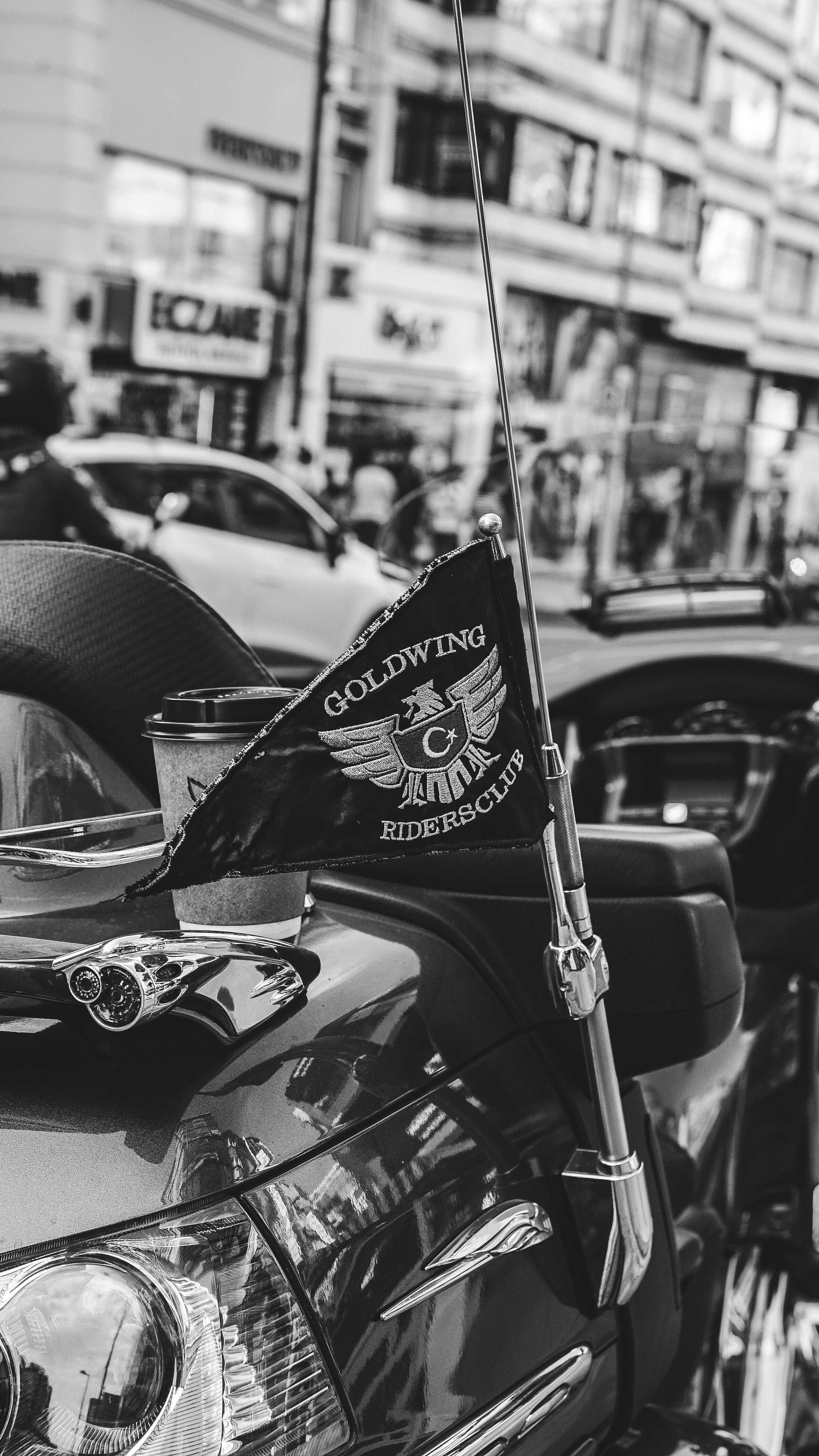 Close-up of a Goldwing Riders Club flag on a motorcycle, showcasing the emblem and a coffee cup in the background. Black and white composition emphasizes the details.