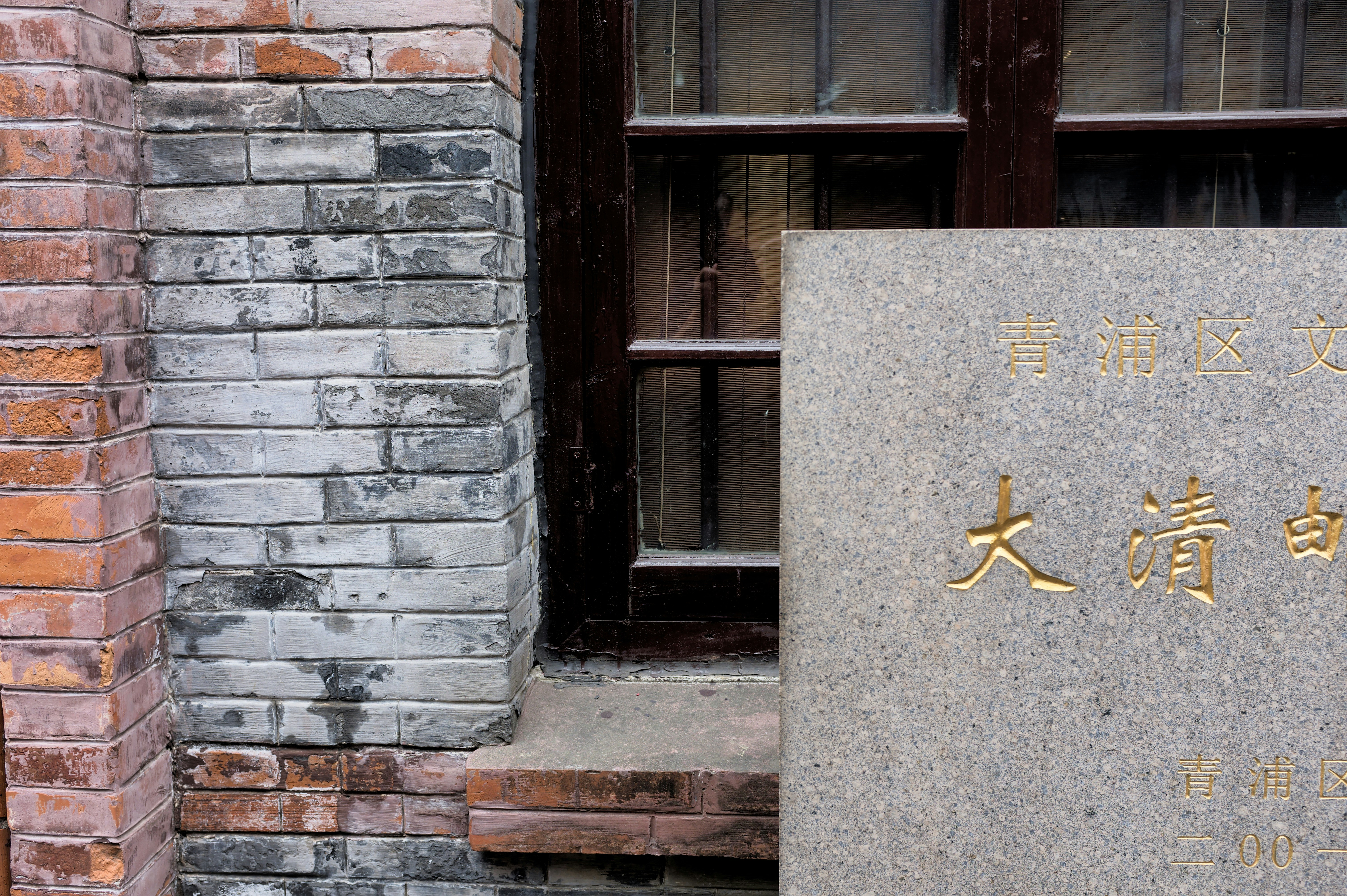 Close-Up of Brick Wall With Window and Stone Sign in Chinese - A weathered brick wall with a window and a stone sign with Chinese characters in gold. The bricks are a mix of red and gray, and the window is framed in dark wood. The sign is made of a rough stone, and the characters are carved into the surface.