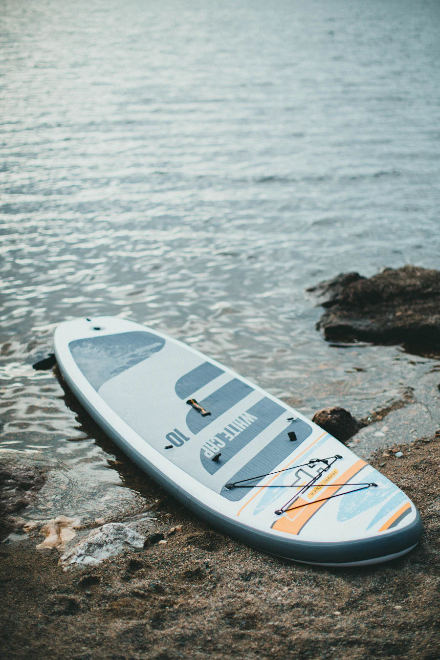 Bright stand-up paddleboard rests on sandy shore with gentle water in the background; board features geometric graphics and leans toward rocks by the waterline.