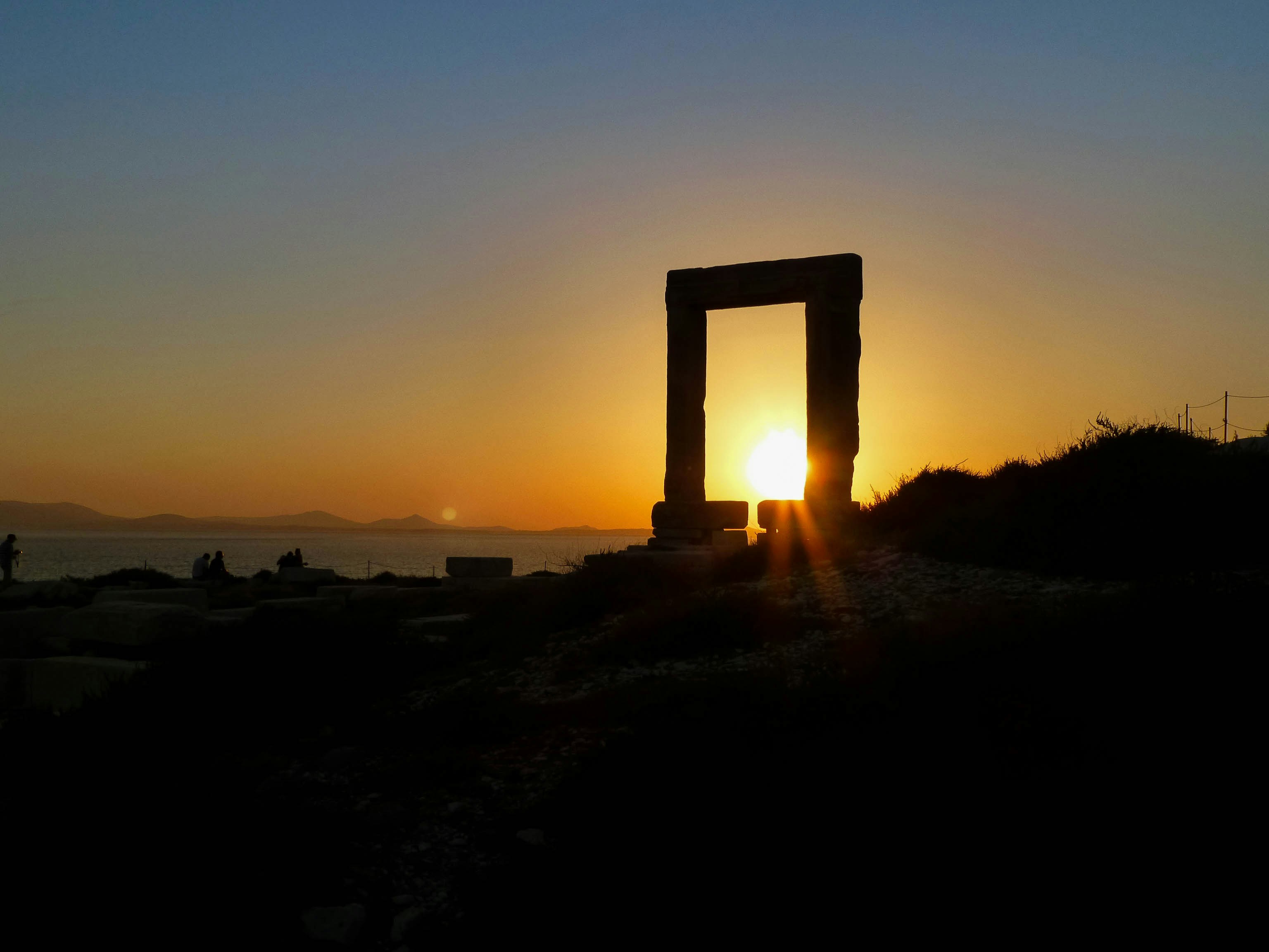 Sunset silhouette of a stone doorway framing the sun above the horizon over a calm sea, with silhouetted dunes in the foreground.