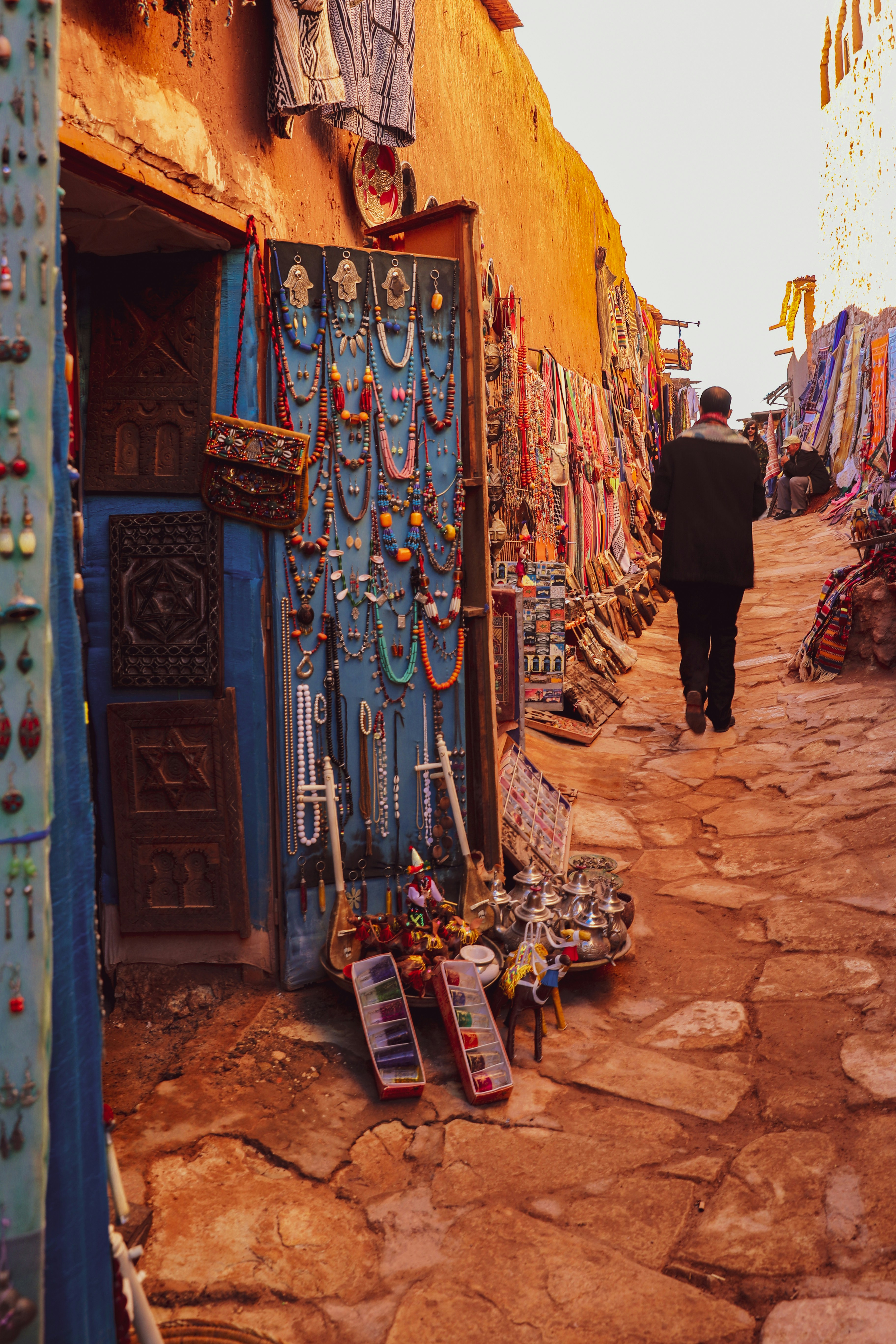 Narrow alleyway adorned with colorful jewelry and trinkets, leading to a figure walking away. The rich textures and hues create an inviting atmosphere.