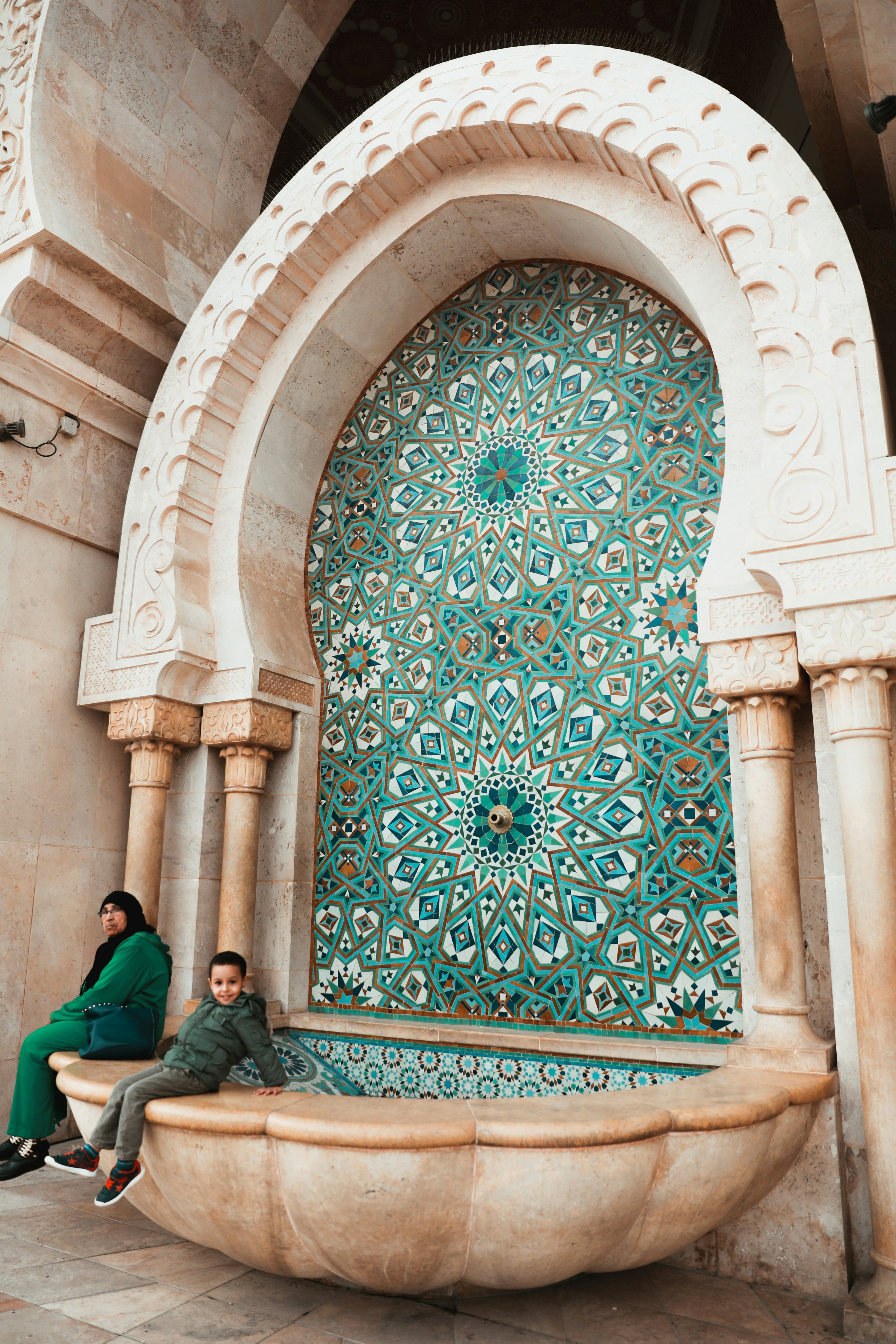 Intricate tiled wall featuring geometric patterns in a serene architectural setting, with a woman and child seated nearby.