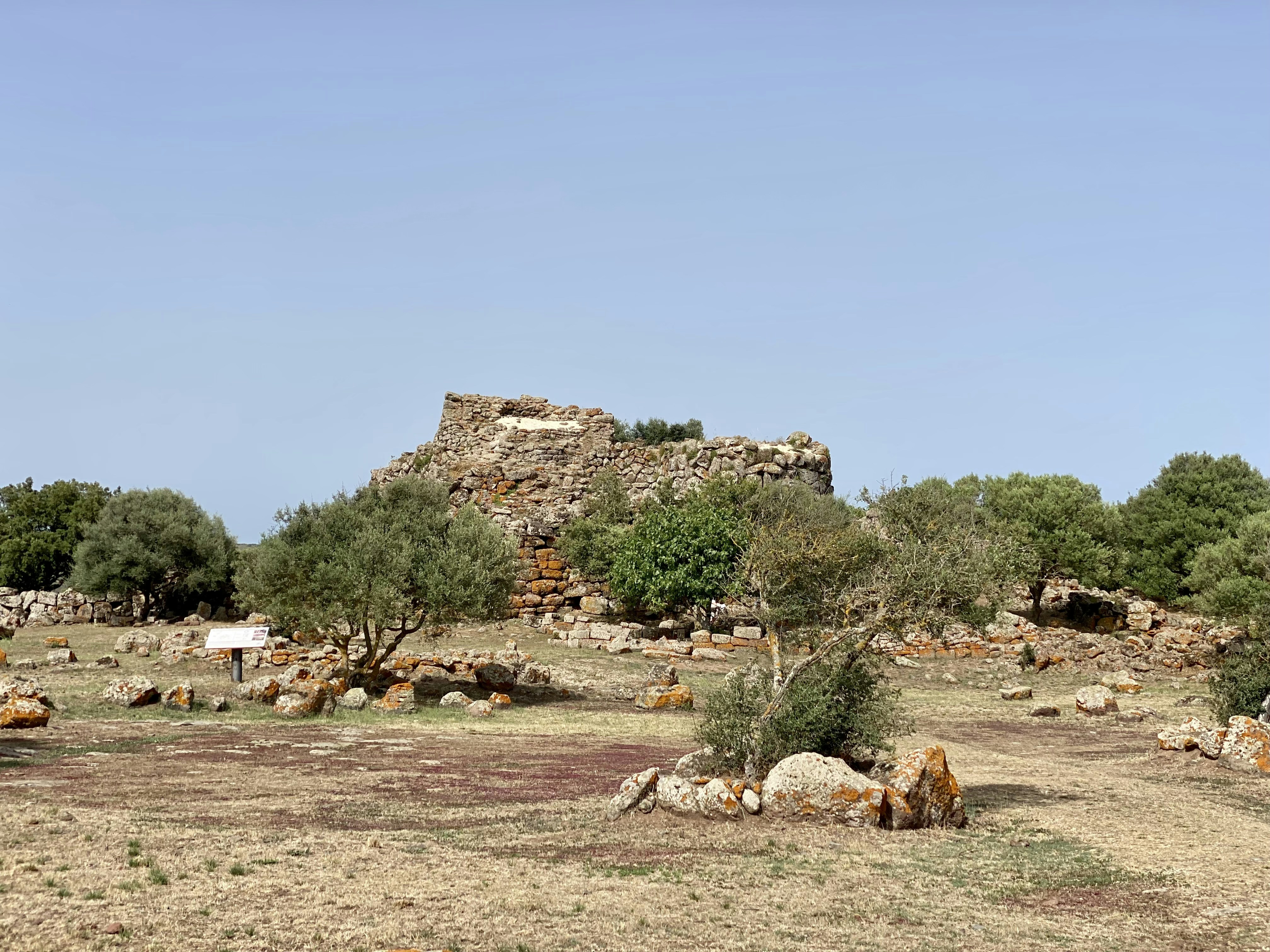 Ruined stone fortress surrounded by olive trees and rocky terrain under a clear sky.