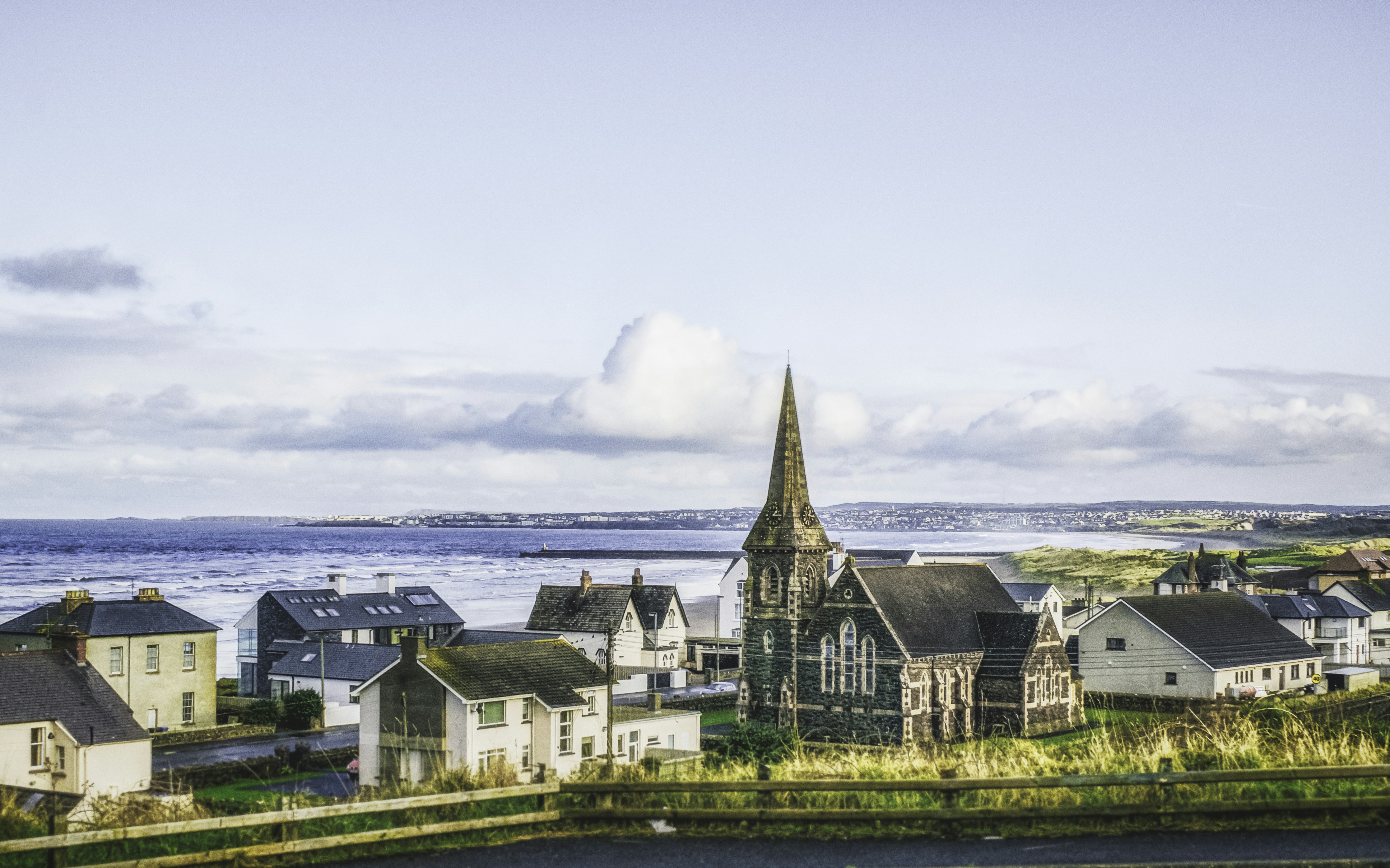 Photograph of a coastal village with a tall church spire and rows of houses along the shoreline, with calm sea in the distance.