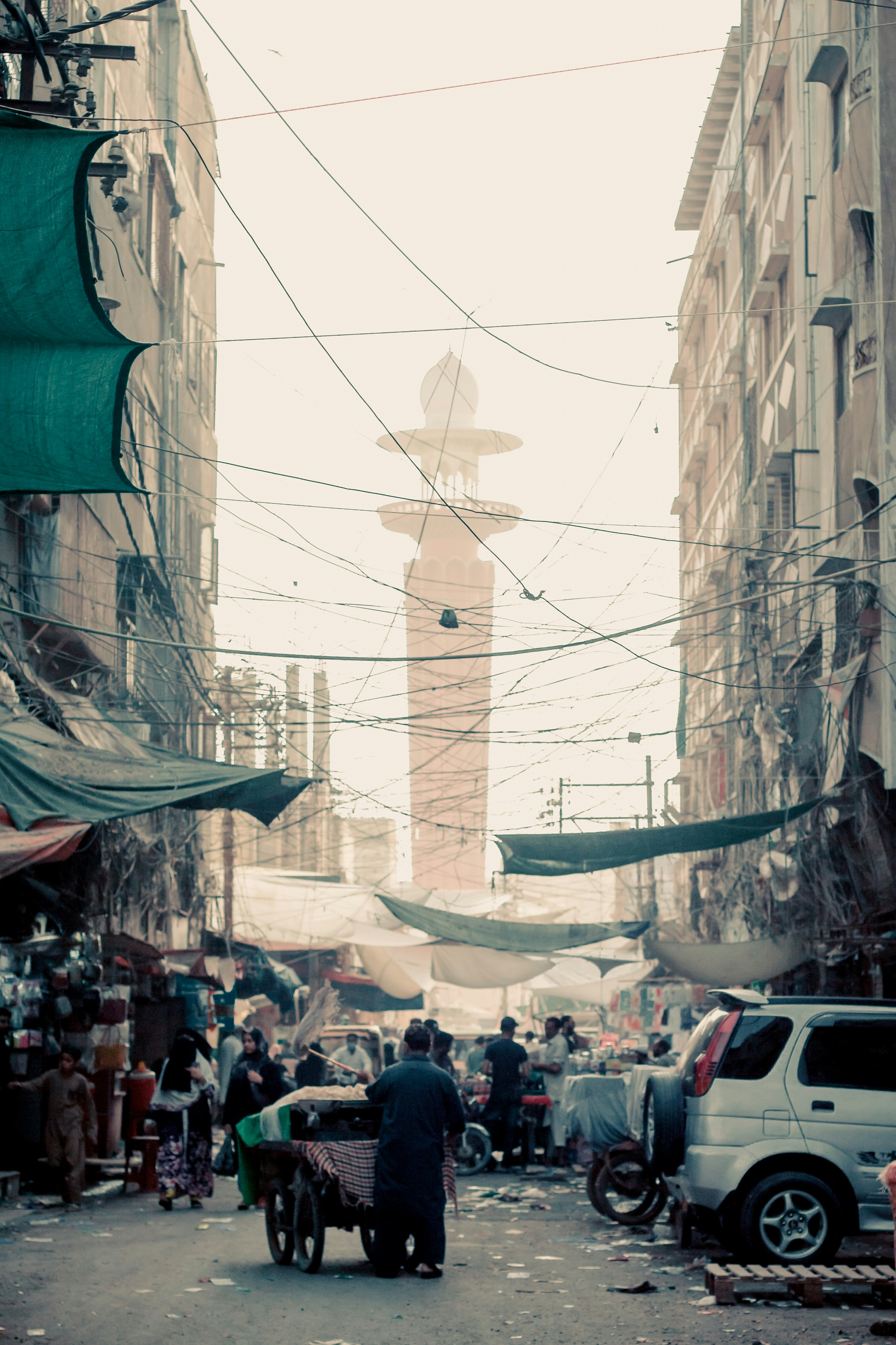 Vibrant market scene filled with vendors and shoppers, featuring a prominent tower in the background amidst tangled overhead wires.