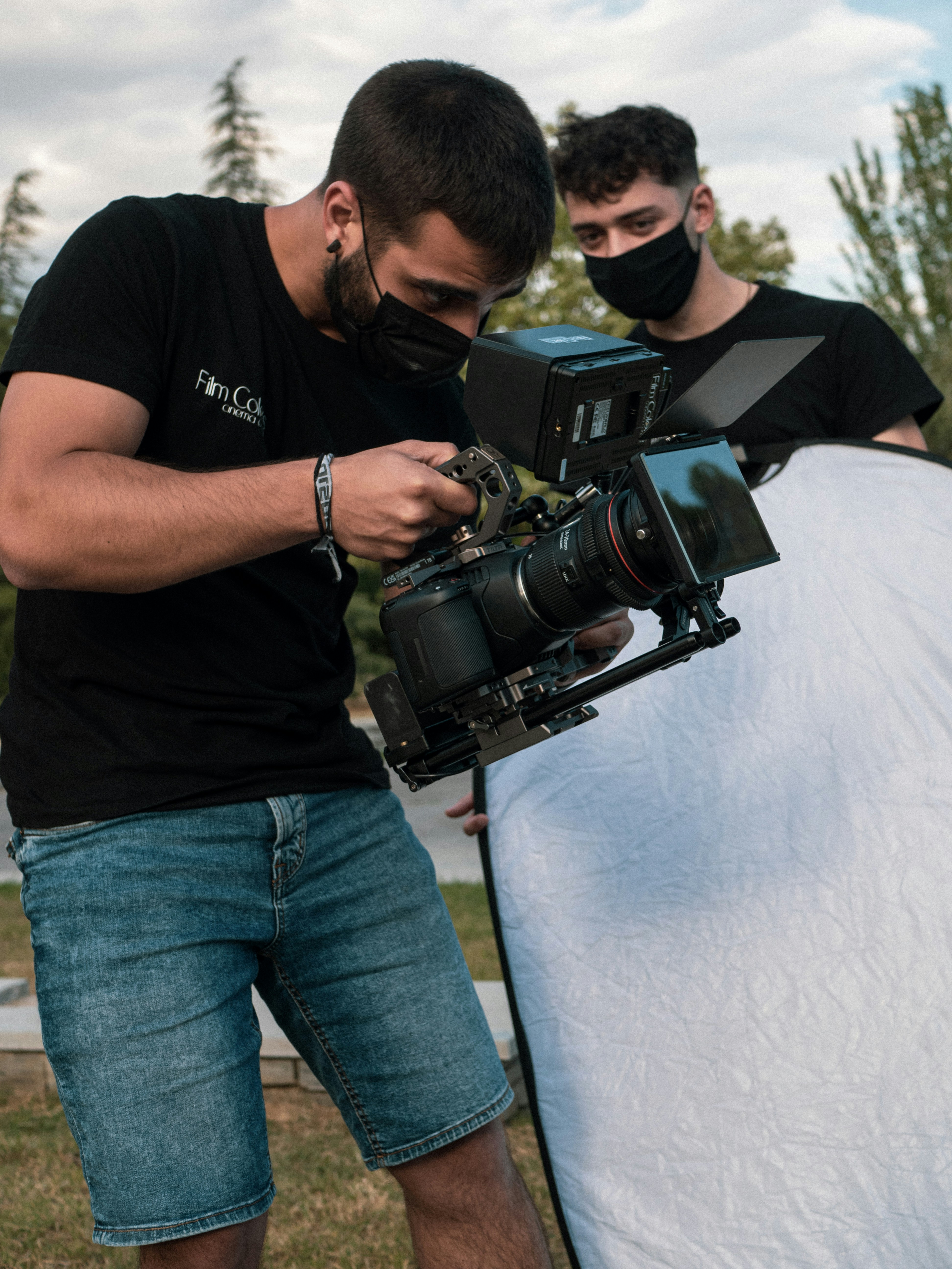 Two filmmakers adjusting equipment outdoors, focusing on a Panasonic DC-G90 camera setup while a reflector is positioned nearby.