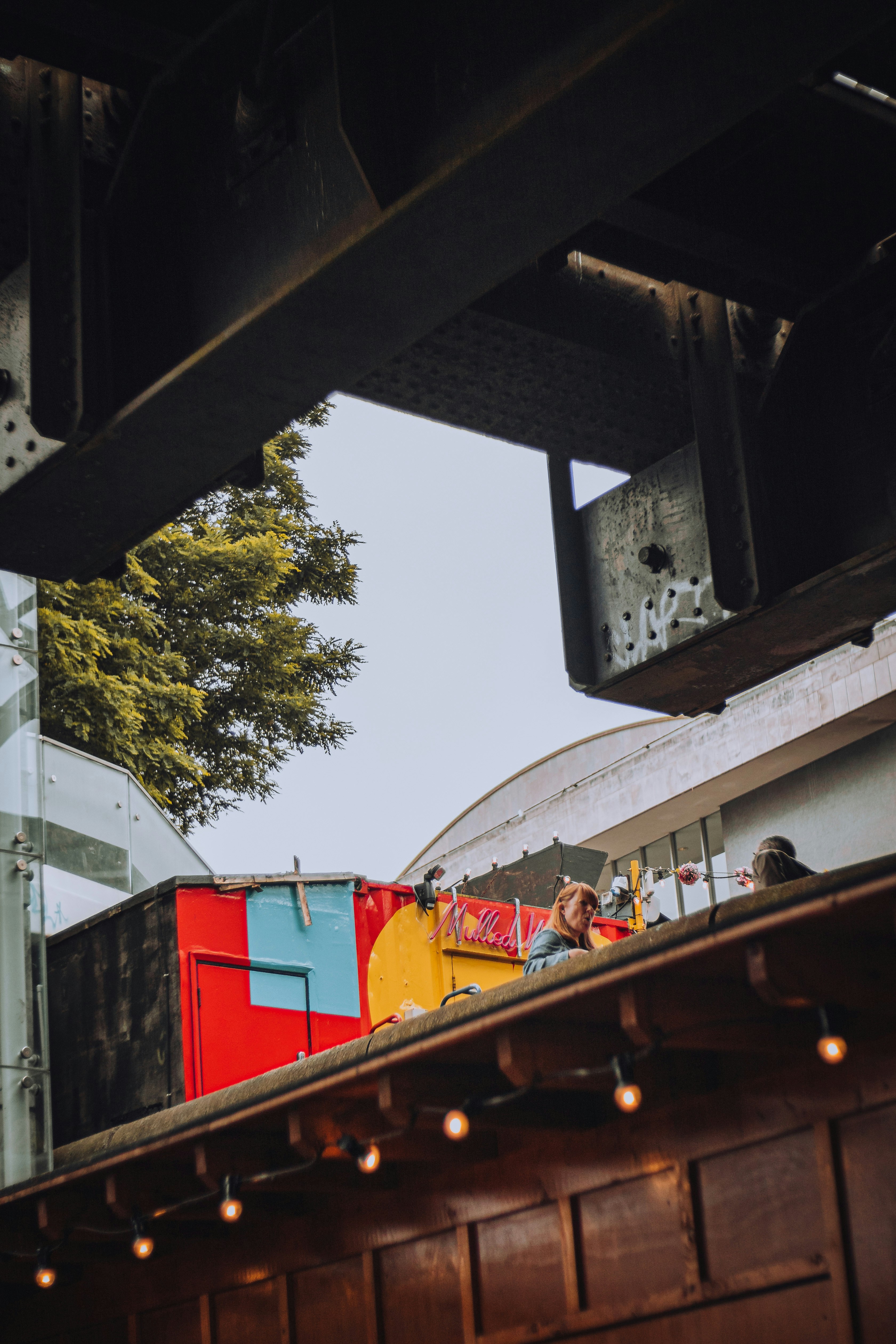 Vibrant food stalls and a lush tree are framed under a metal overpass, showcasing an urban setting filled with life and color.