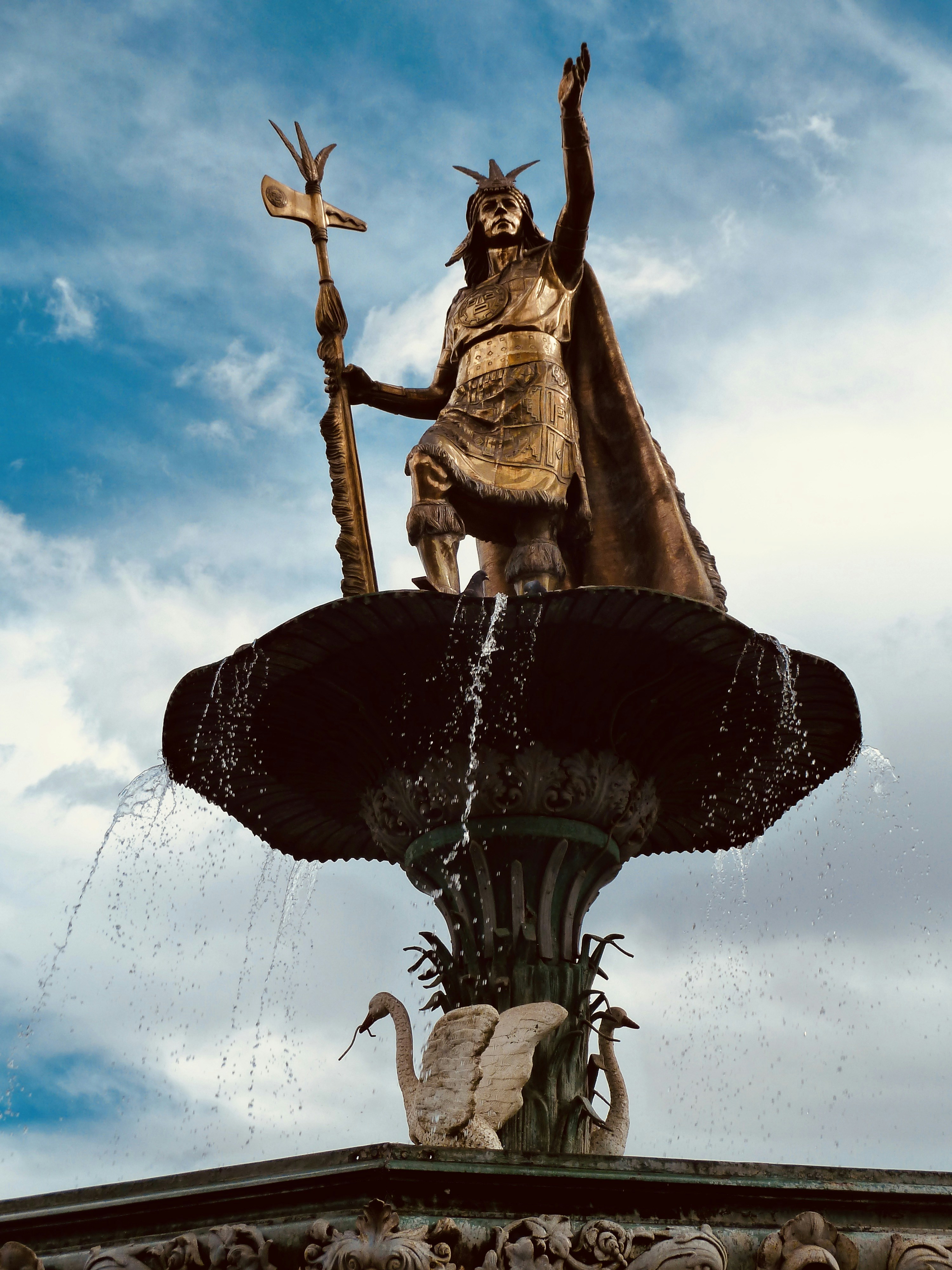 Bronze statue of a historical figure atop a fountain, surrounded by flowing water and intricate sculptures. The sky showcases a dramatic blue backdrop.