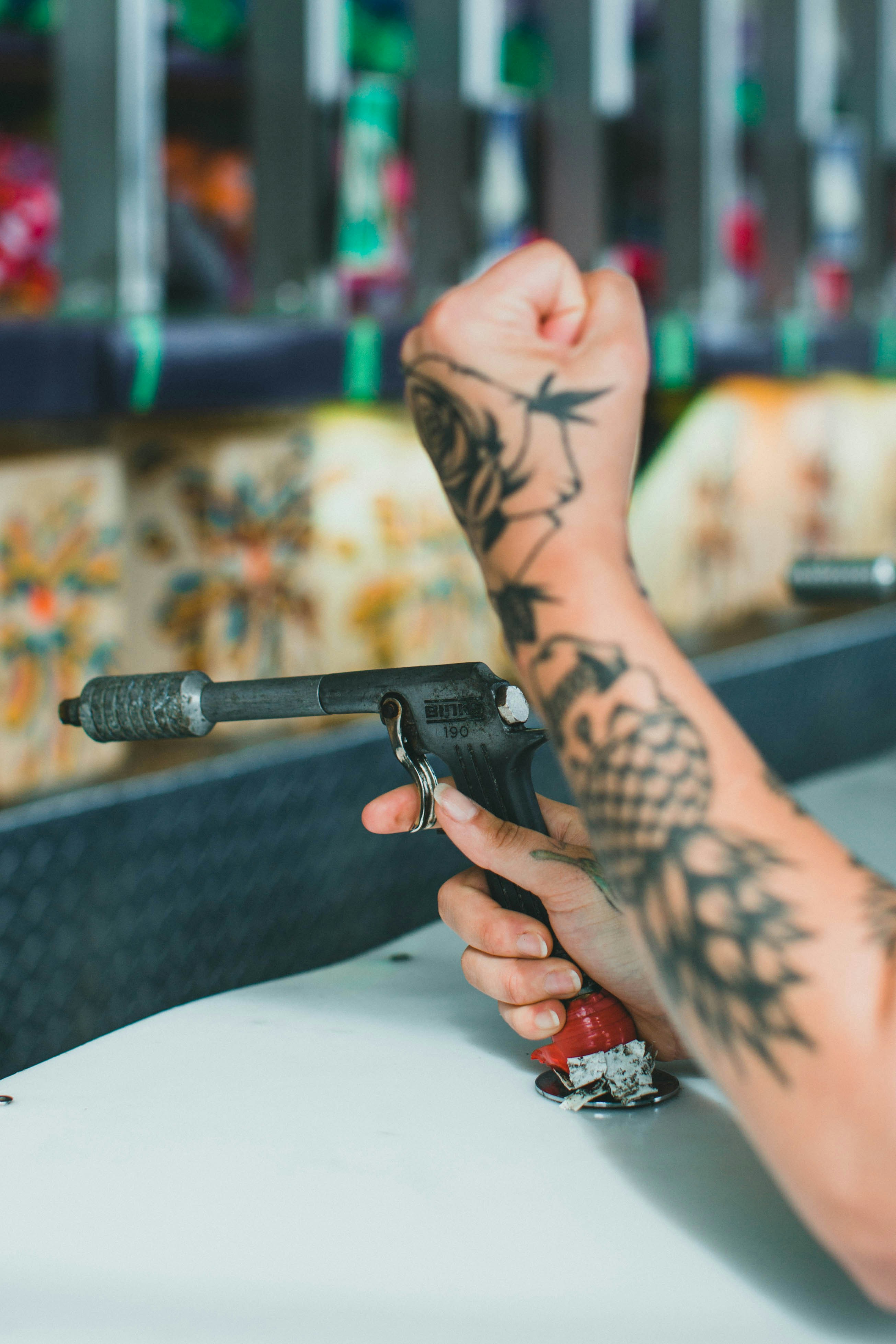 Close-up photograph of a tattooed forearm gripping a spray gun against a graffiti-filled skate park backdrop.