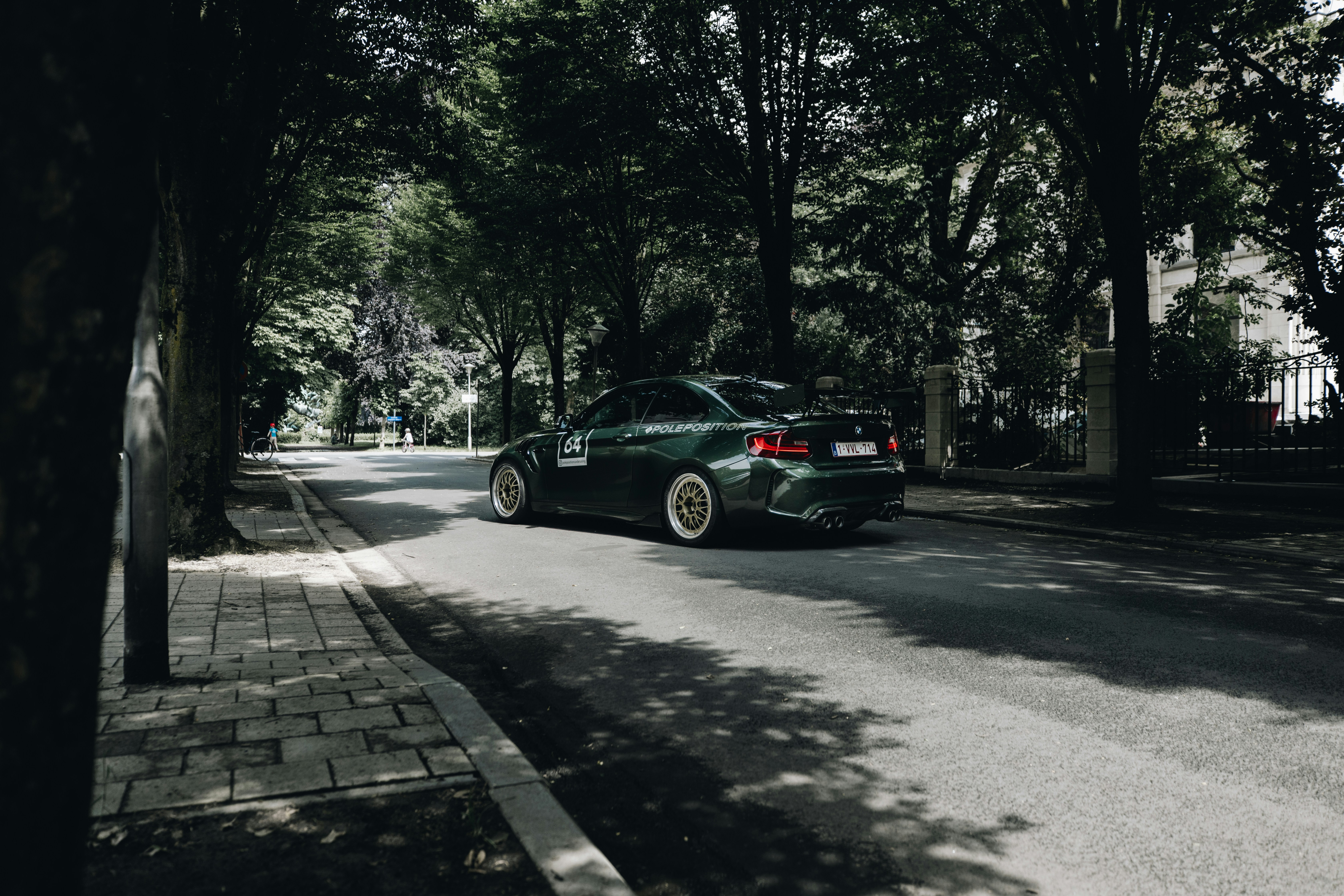 A modified car with distinctive wheels navigates a tree-lined street, showcasing urban automotive culture. The scene captures a blend of nature and machinery.