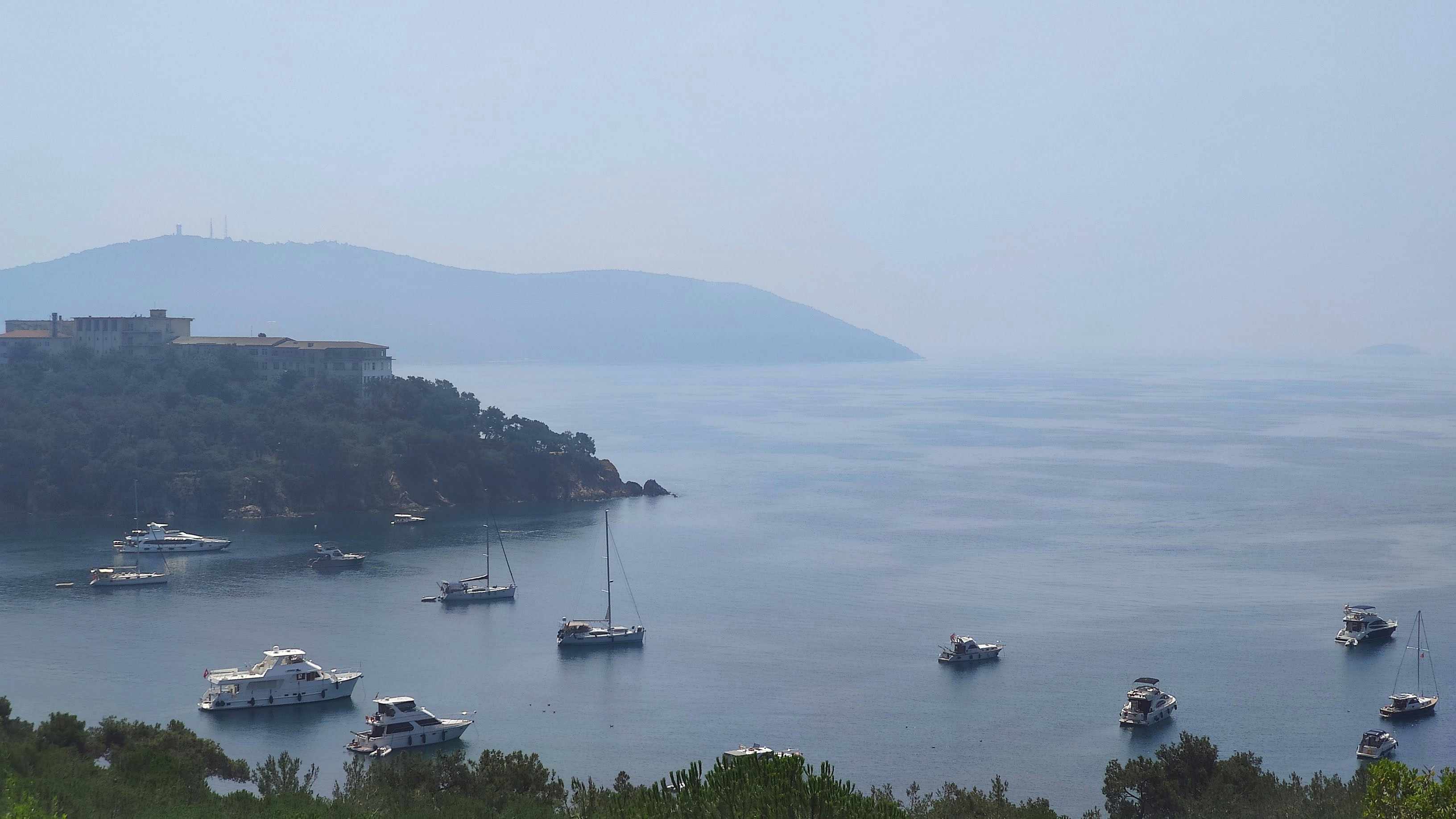 A tranquil harbor scene featuring several boats anchored in calm waters, set against a hazy backdrop of distant hills and lush greenery.