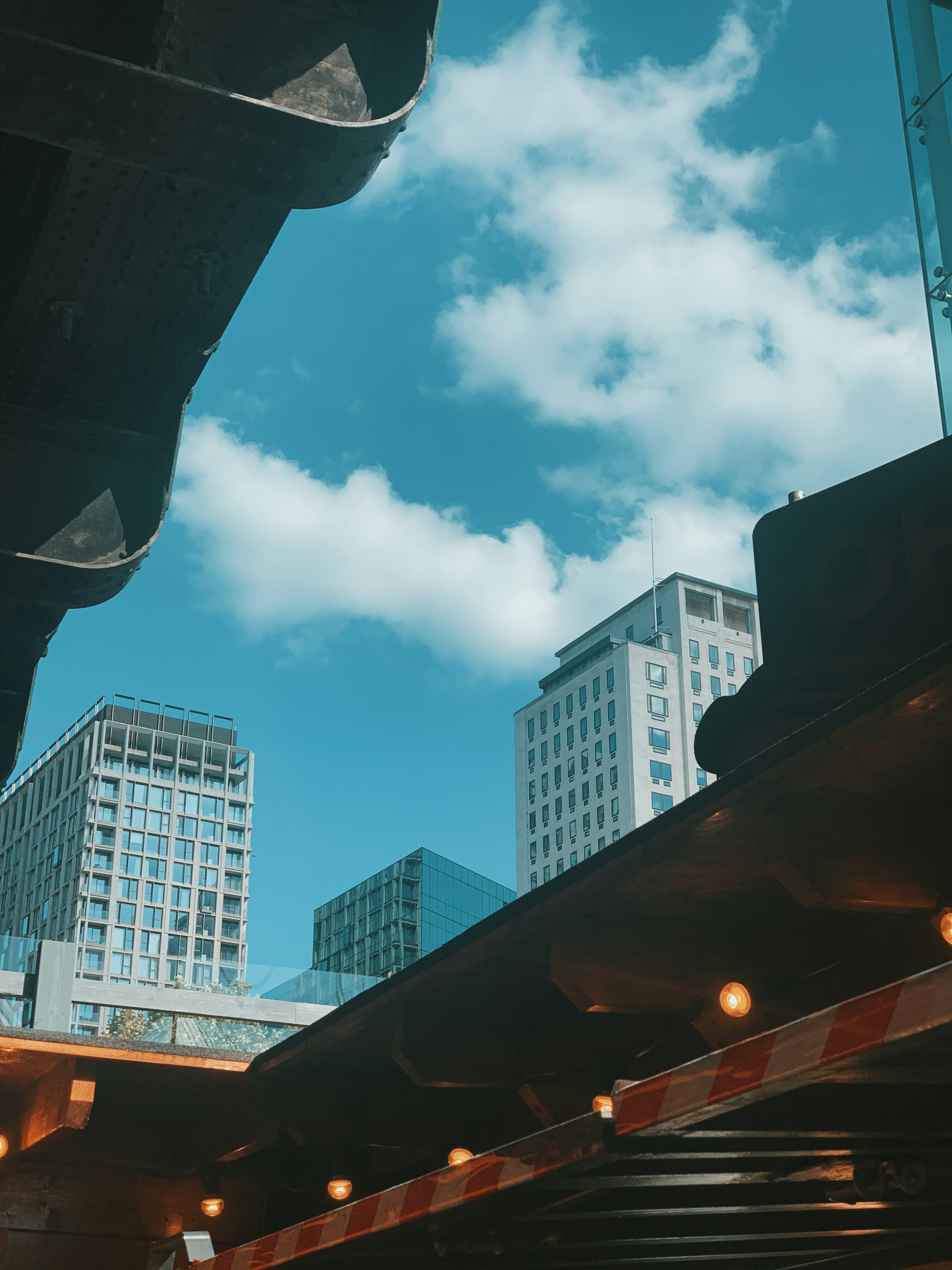 A view of modern skyscrapers under a bright blue sky, framed by architectural elements above. Fluffy clouds drift lazily in the background.