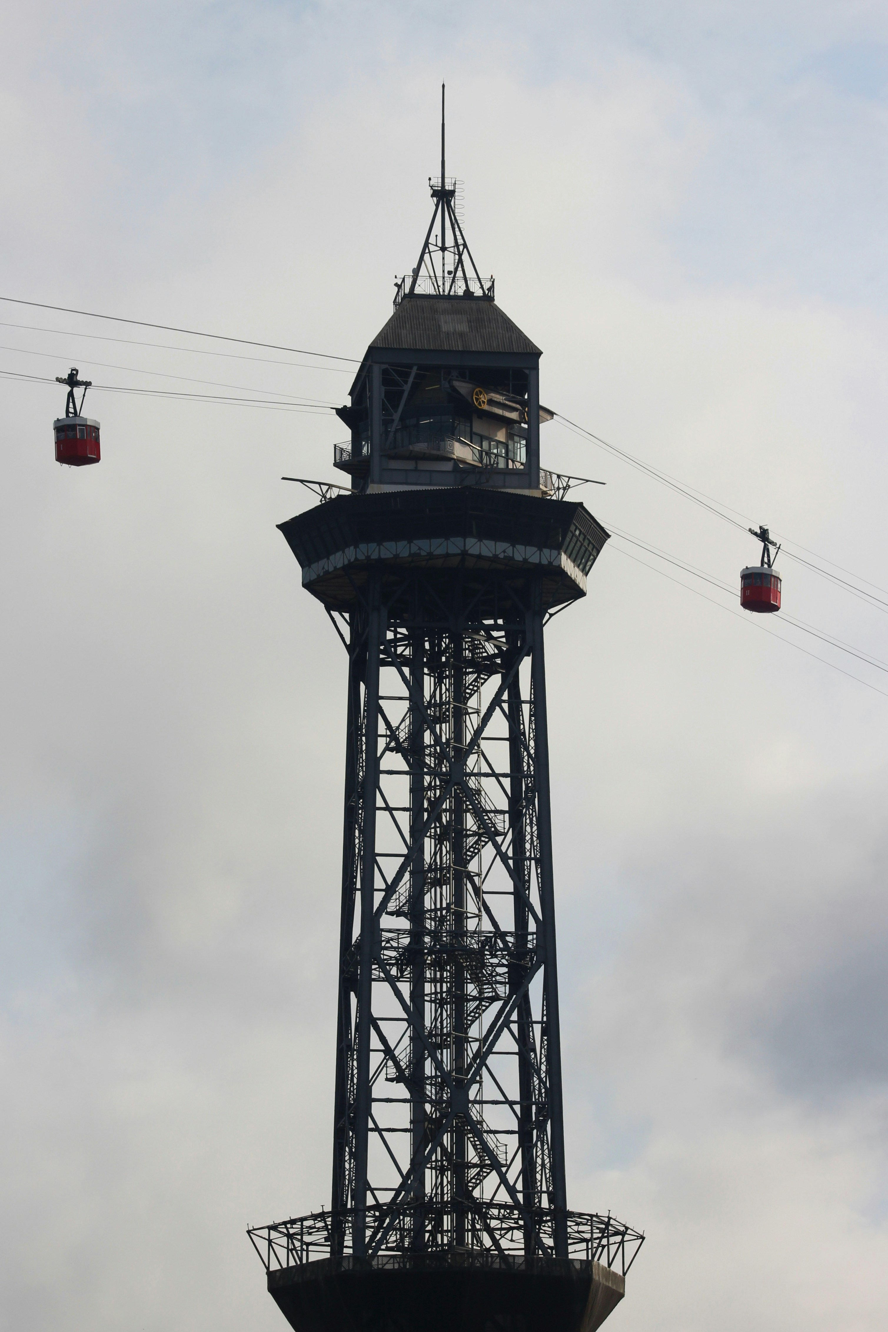 Aerial tramway tower with red gondolas traversing cables against a cloudy sky. The structure showcases engineering elegance and functionality.