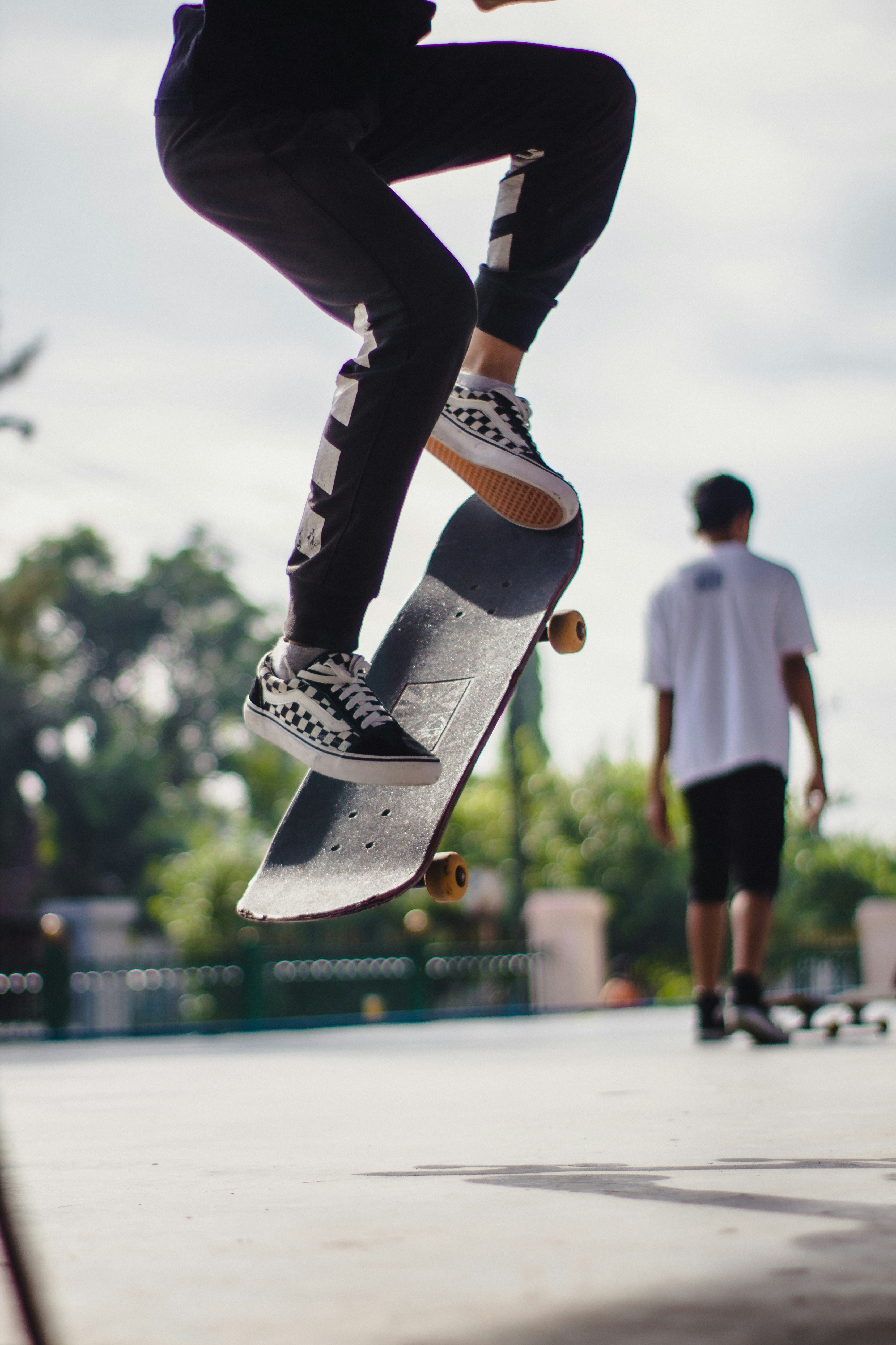Skateboarder performing a jump with dynamic motion, captured in a skate park environment. The background features a second skater and lush greenery.