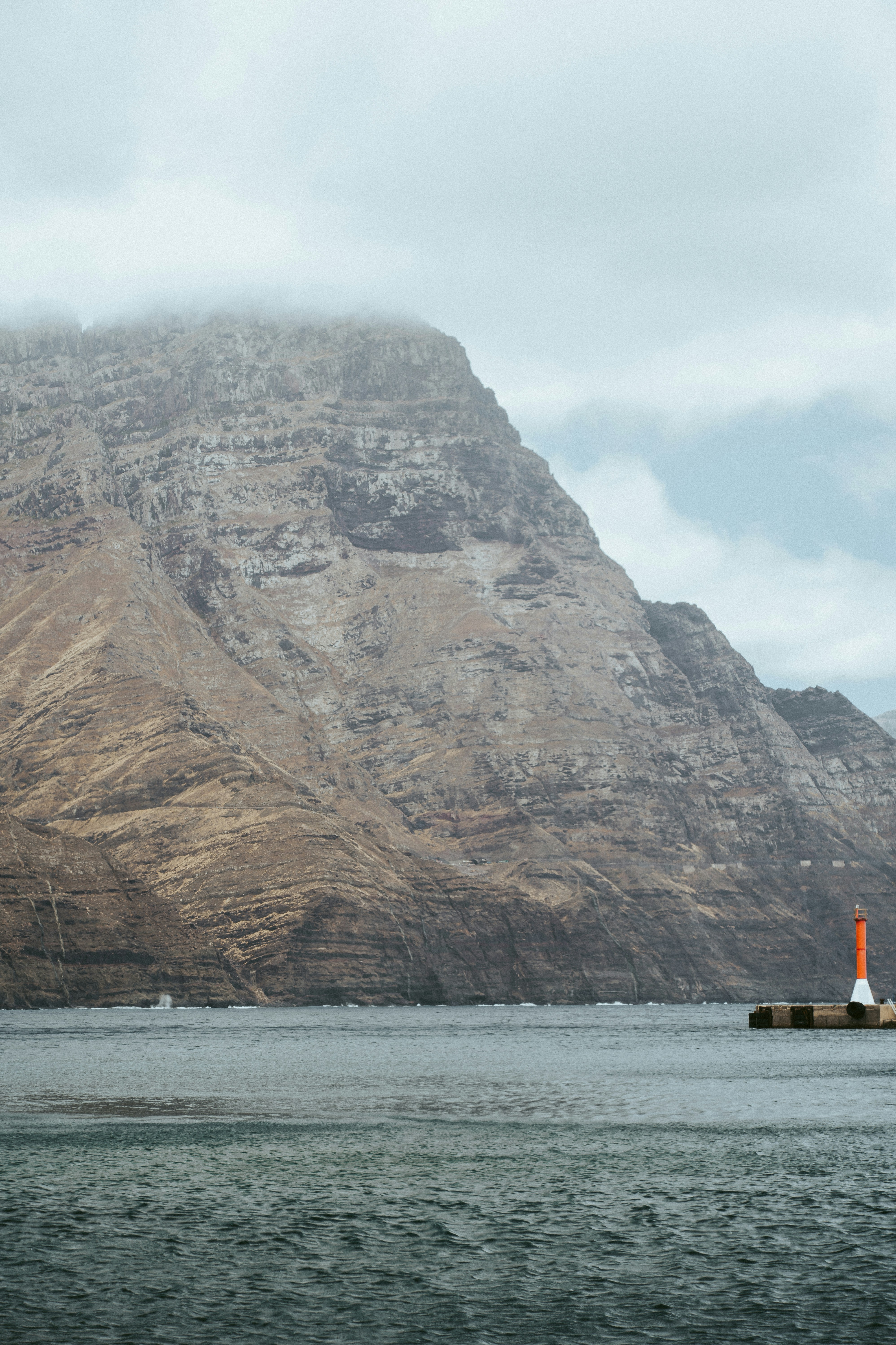 Lighthouse standing on a breakwater in a tranquil harbor, framed by towering, rugged mountains under a cloudy sky.