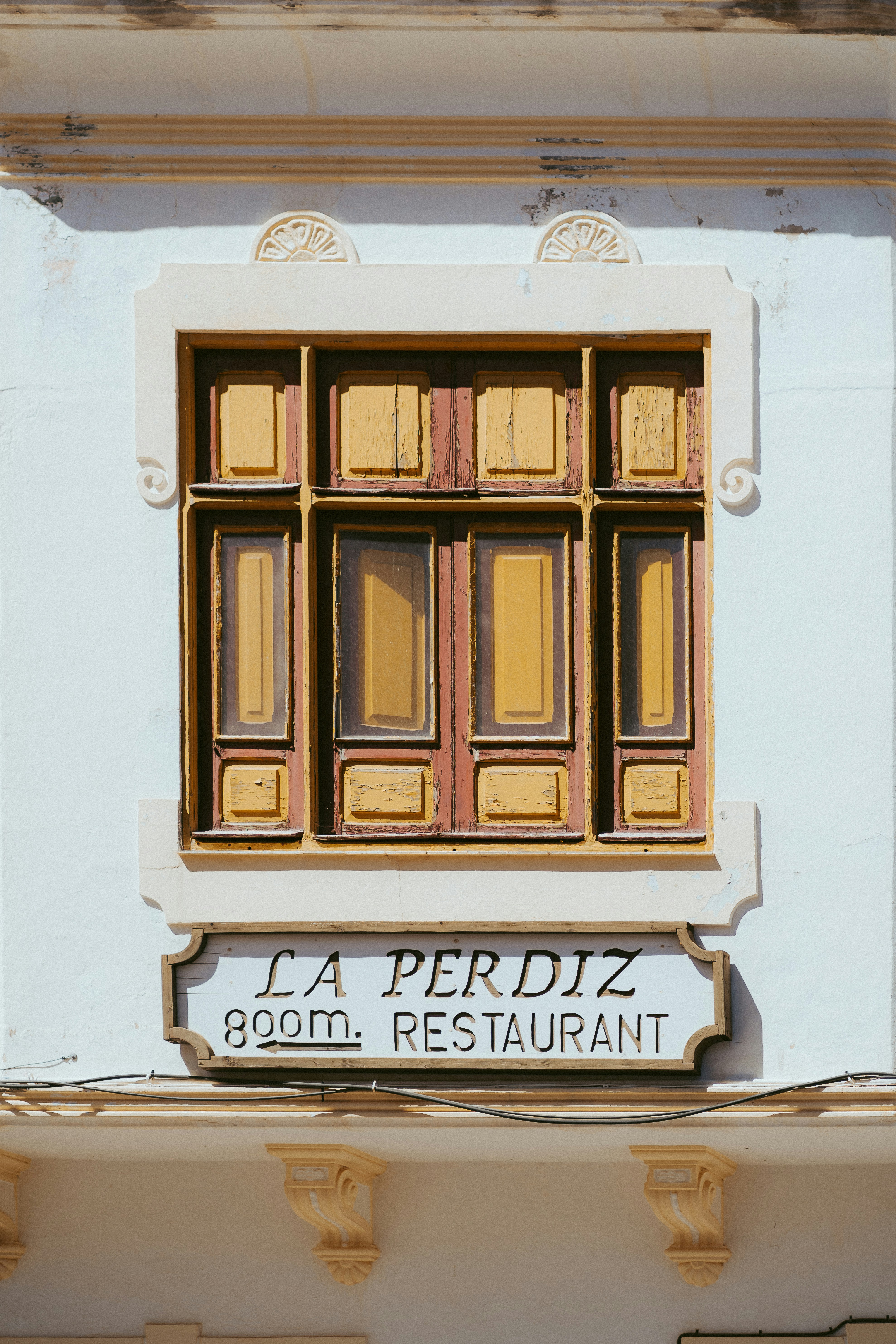 Colorful wooden window with intricate detailing above a restaurant sign in a sunlit setting.