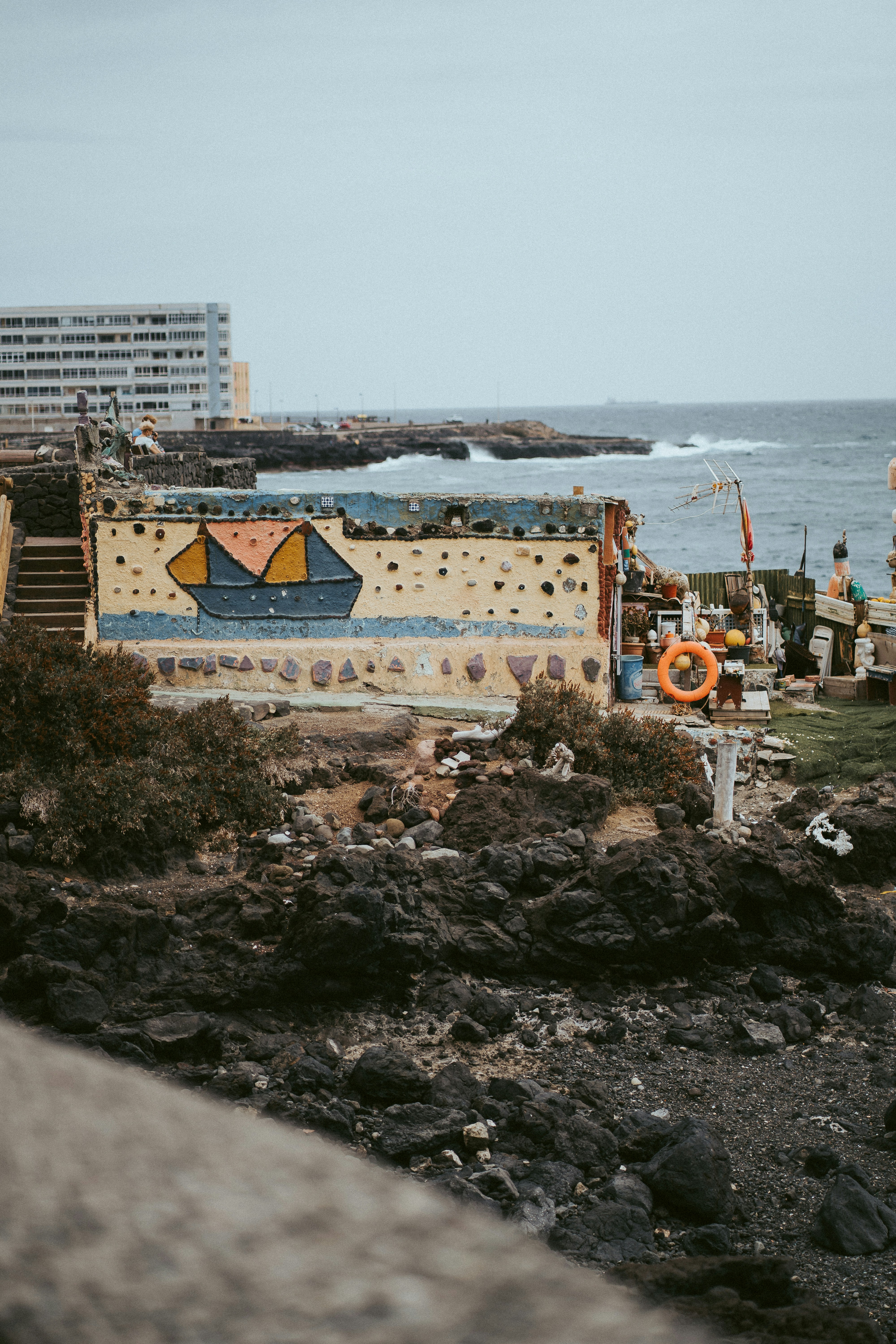 Colorful mural of a boat on a textured wall by the sea, surrounded by rocky terrain and a glimpse of coastal life.