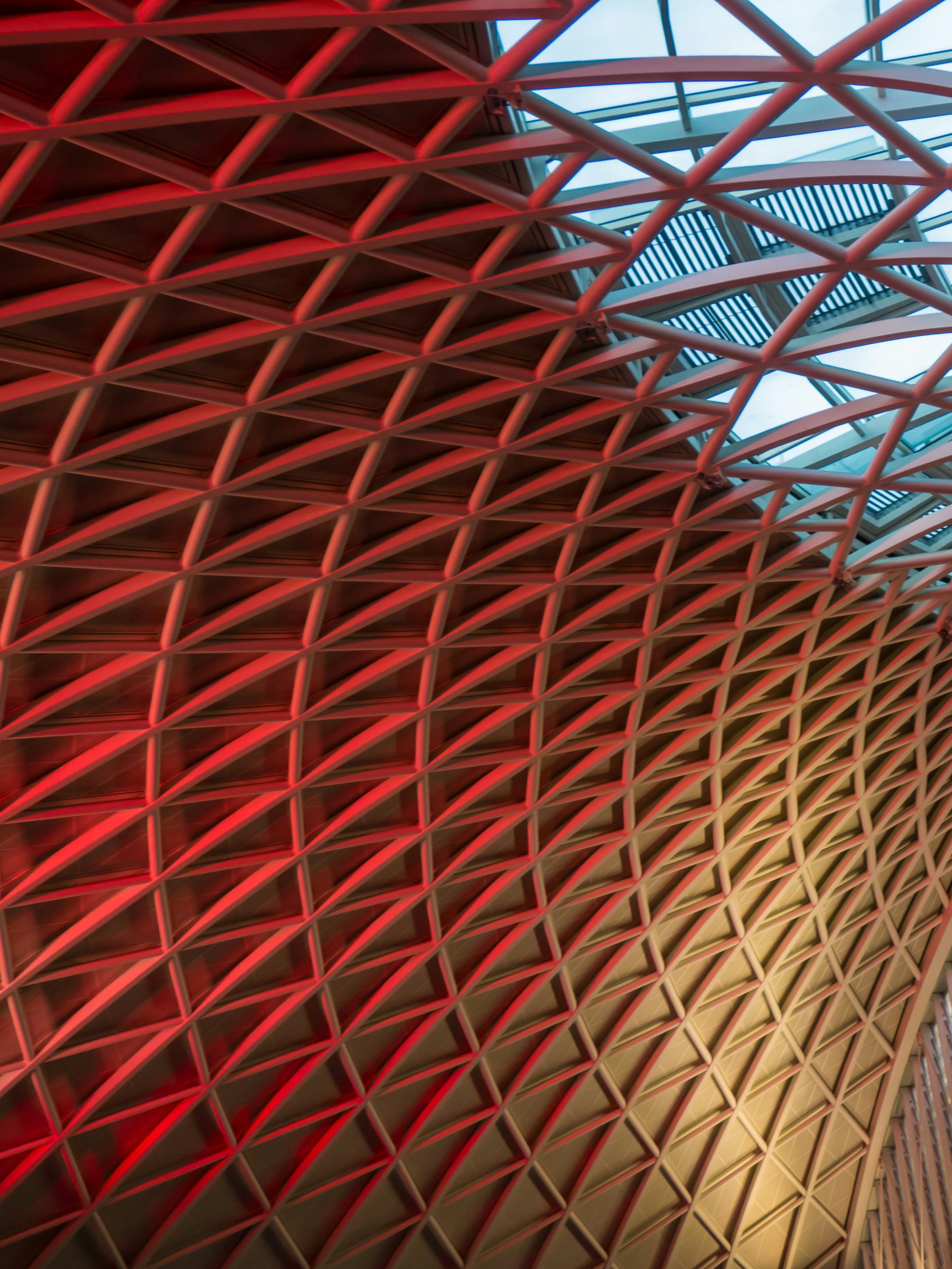Architectural photograph of a red lattice dome curving across the frame, with blue sky visible through the gaps. The repeating diamond pattern creates depth and dramatic light.