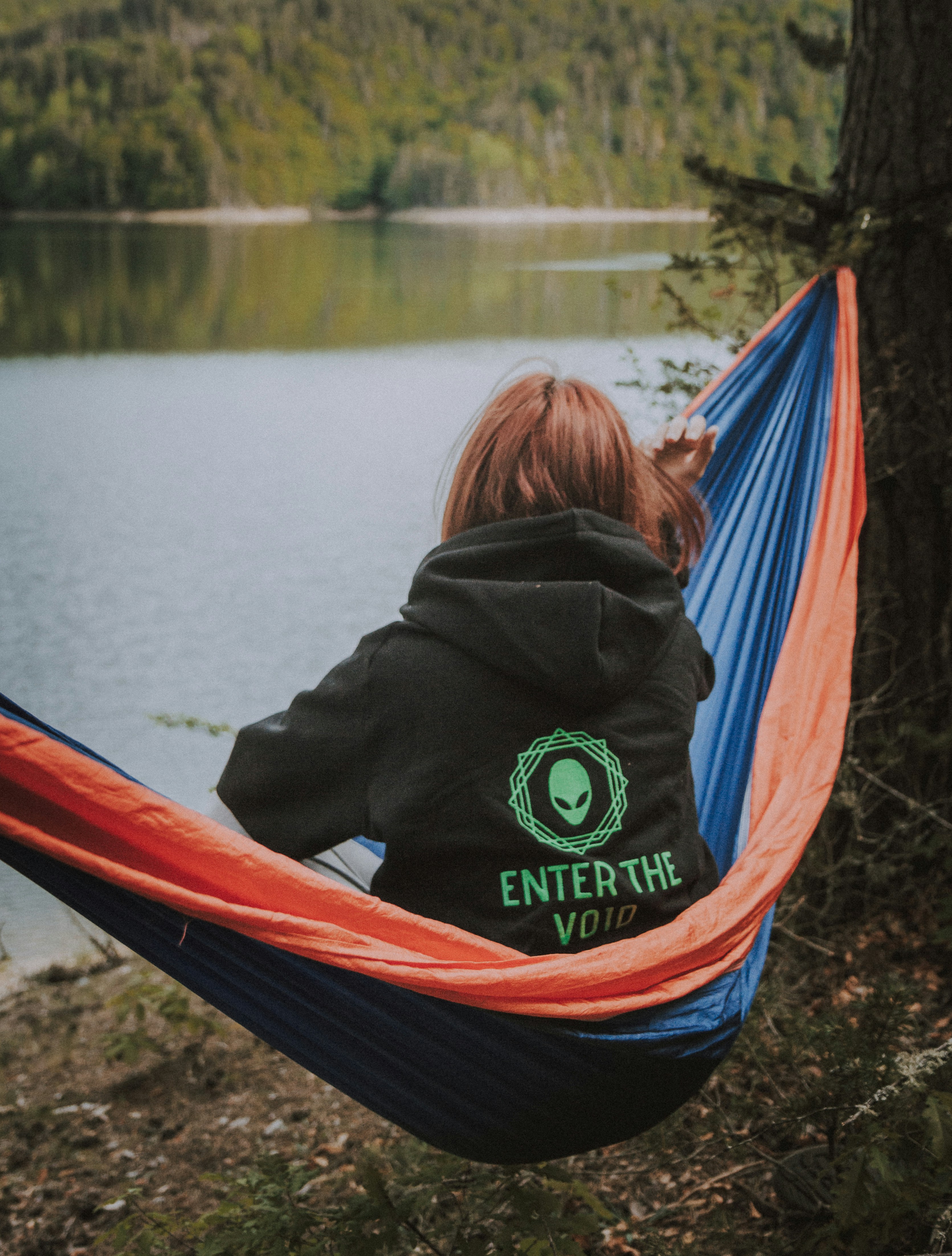 A redhead girl with a hoodie that says "Enter the Void", enjoying the view of the lake while hanging on a hammock between two trees