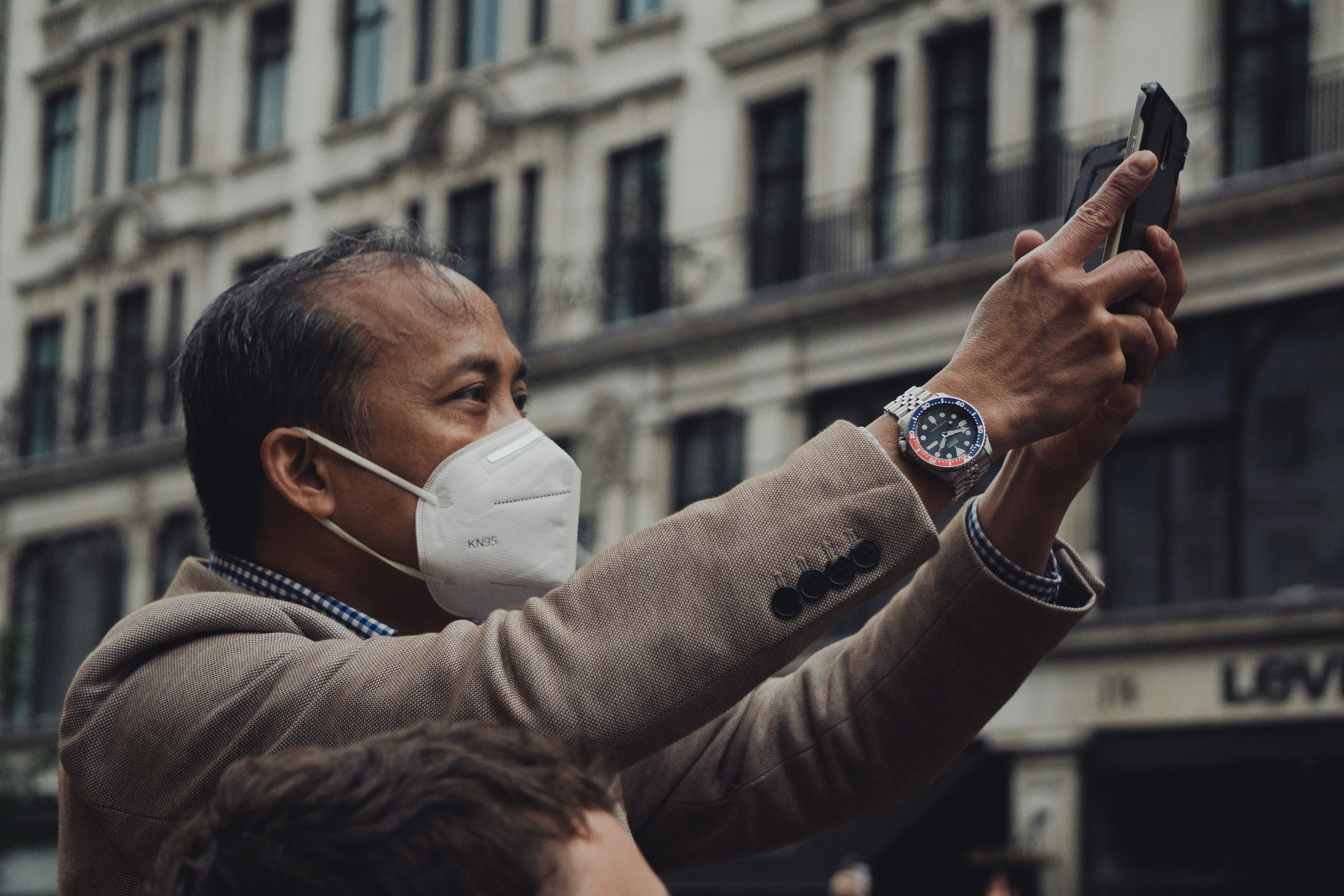 A man wearing a mask captures a moment on his smartphone in a bustling city environment, showcasing the blend of technology and daily life.
