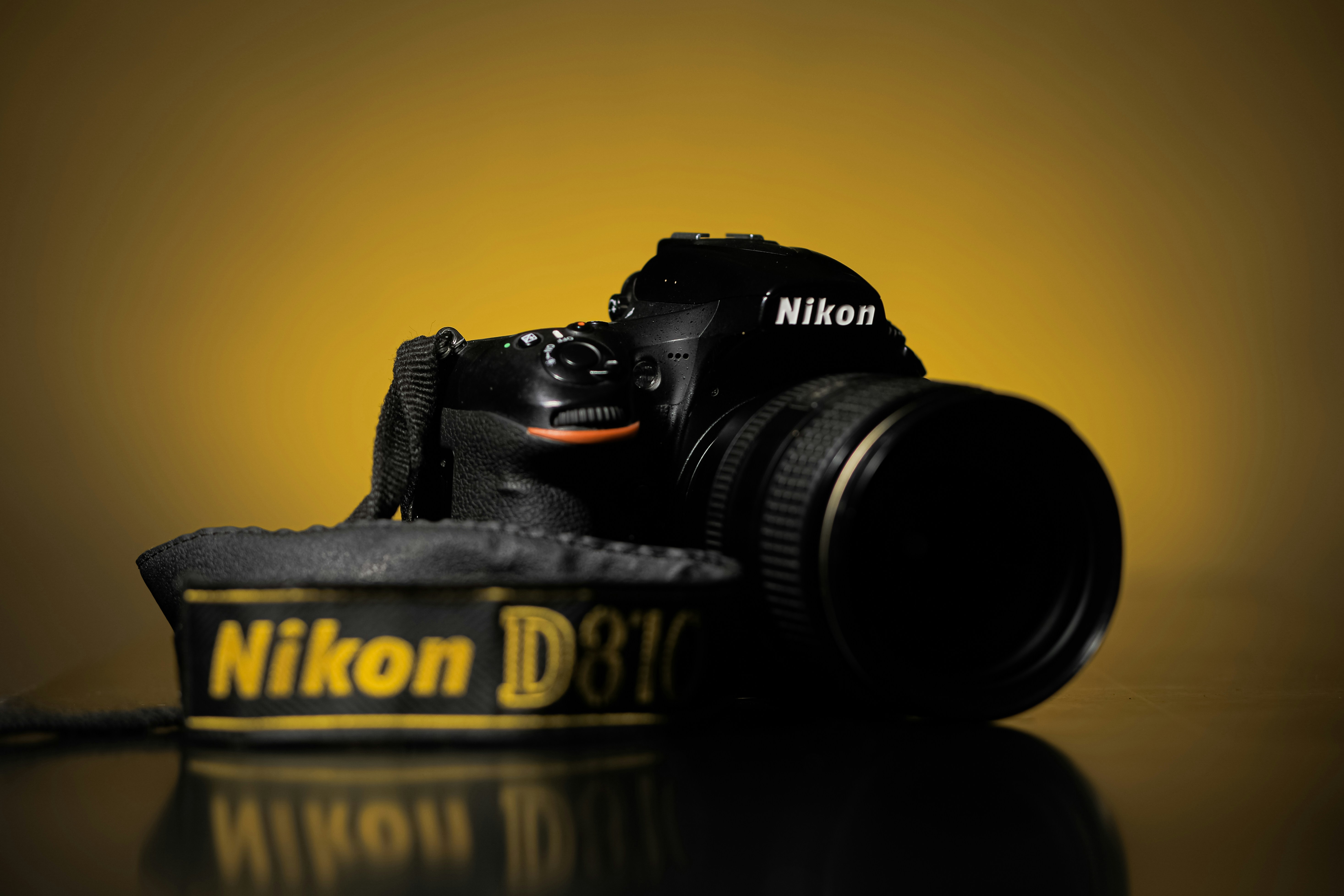 Nikon D810 camera positioned on a reflective surface with a warm yellow background, showcasing its sleek design and branding.