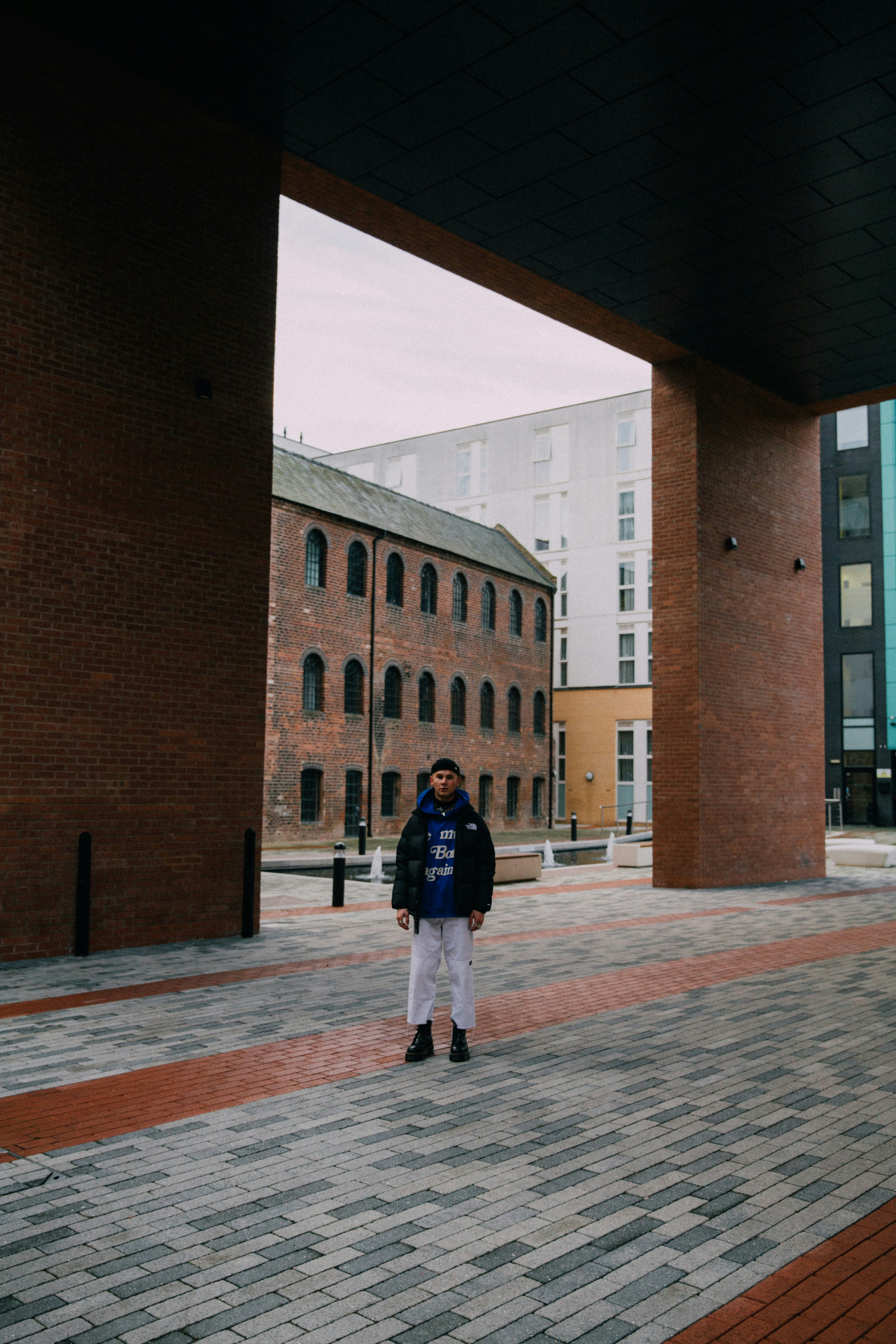 A person stands confidently in a contemporary urban setting, framed by striking brick architecture and modern buildings. The scene highlights the blend of old and new in city design.
