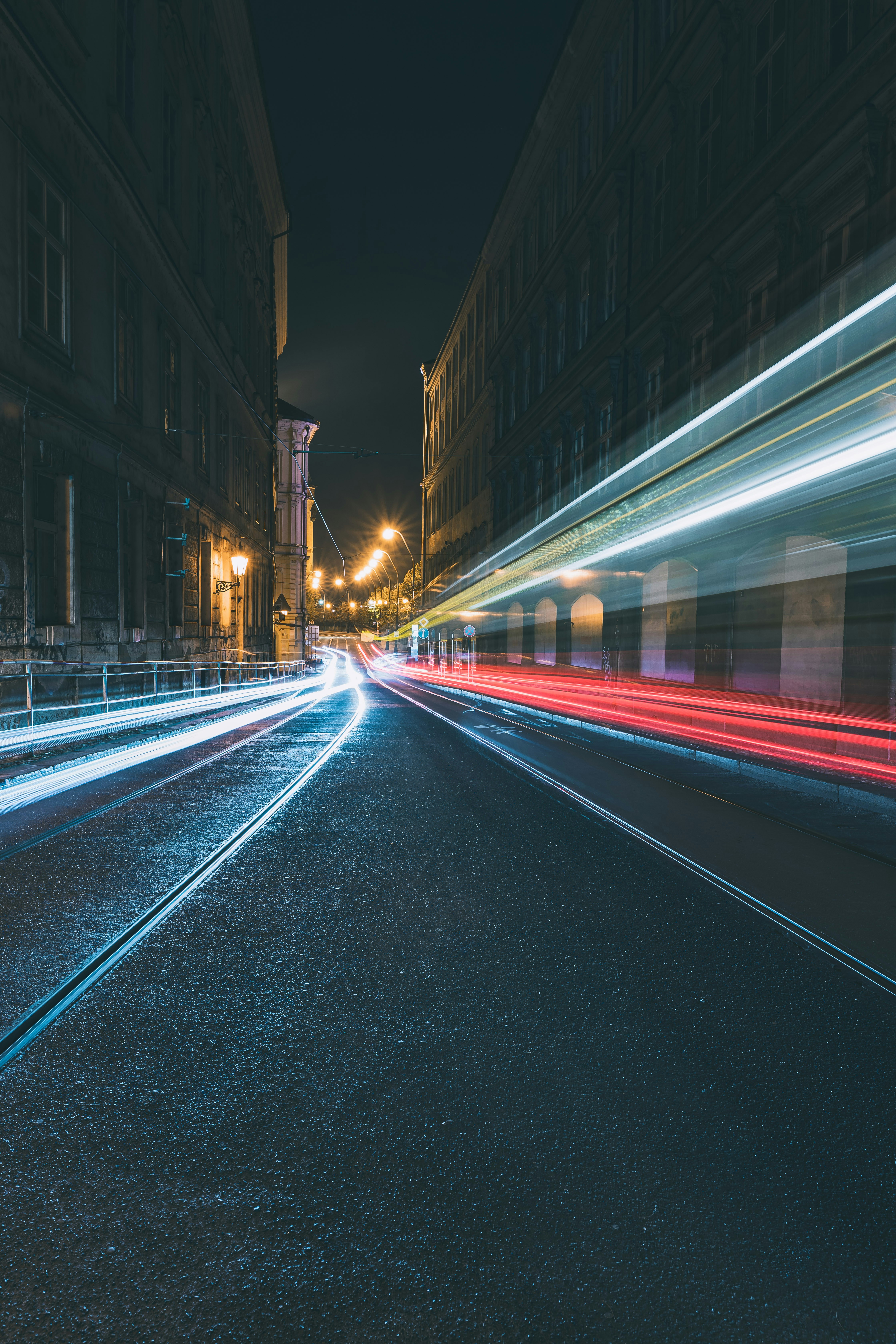 Dynamic light trails weave through a narrow city street, illuminated by street lamps and architectural details. The scene captures the essence of urban movement at night.