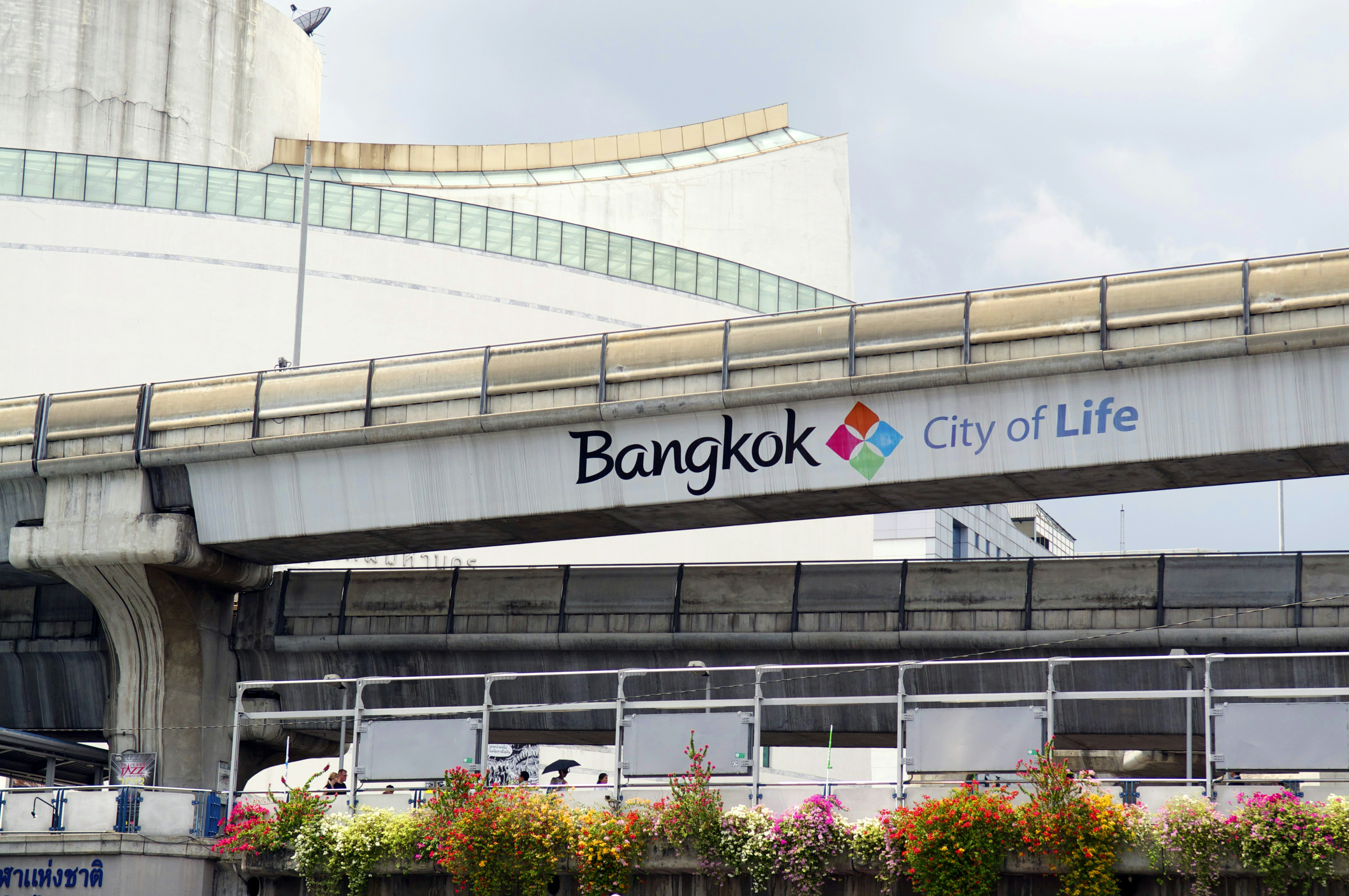 Skytrain track with 'Bangkok City of Life' slogan and colorful flowers below under a cloudy sky.