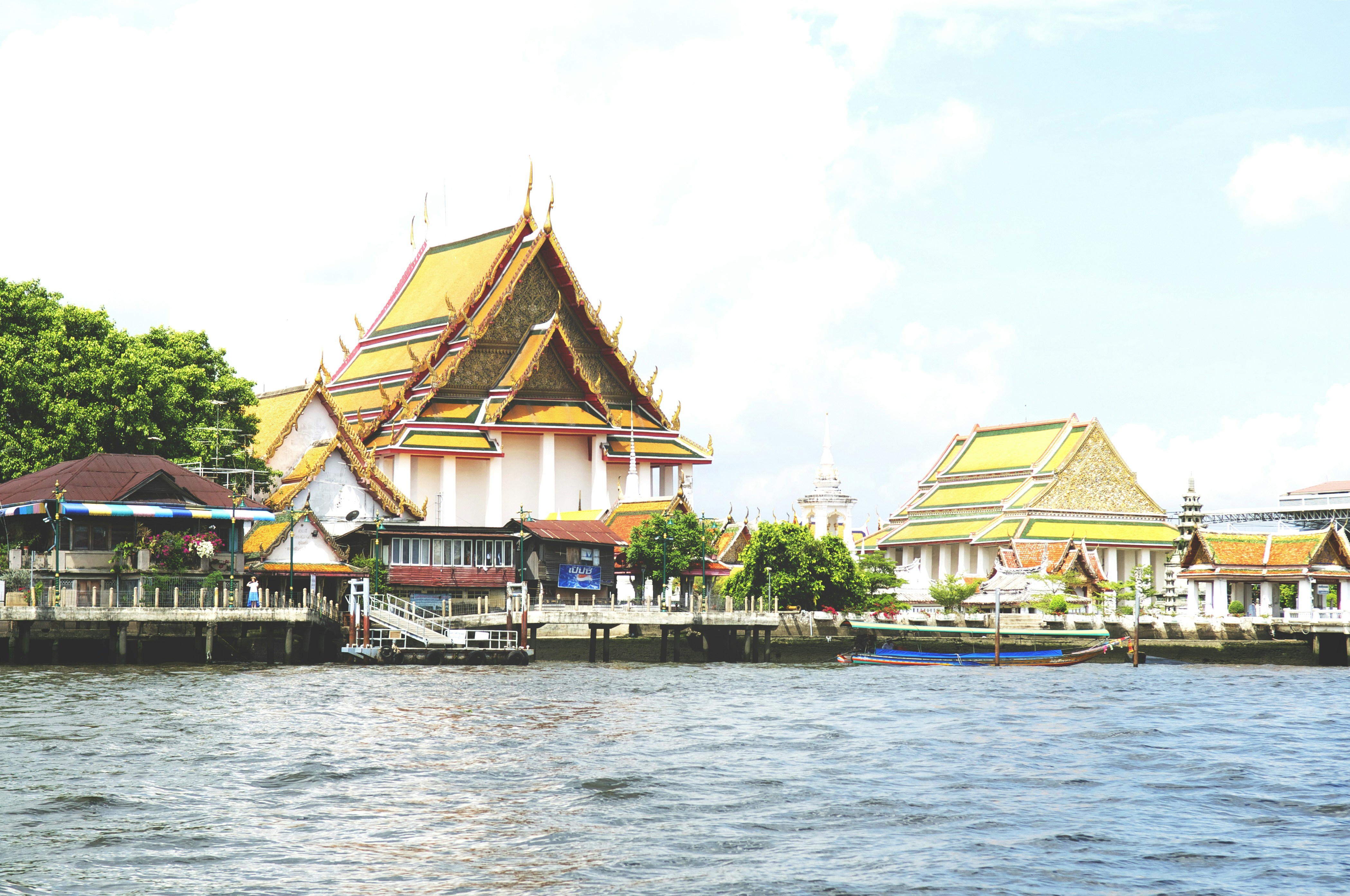 Traditional Thai architecture with ornate rooftops along the Chao Phraya River under a bright sky.