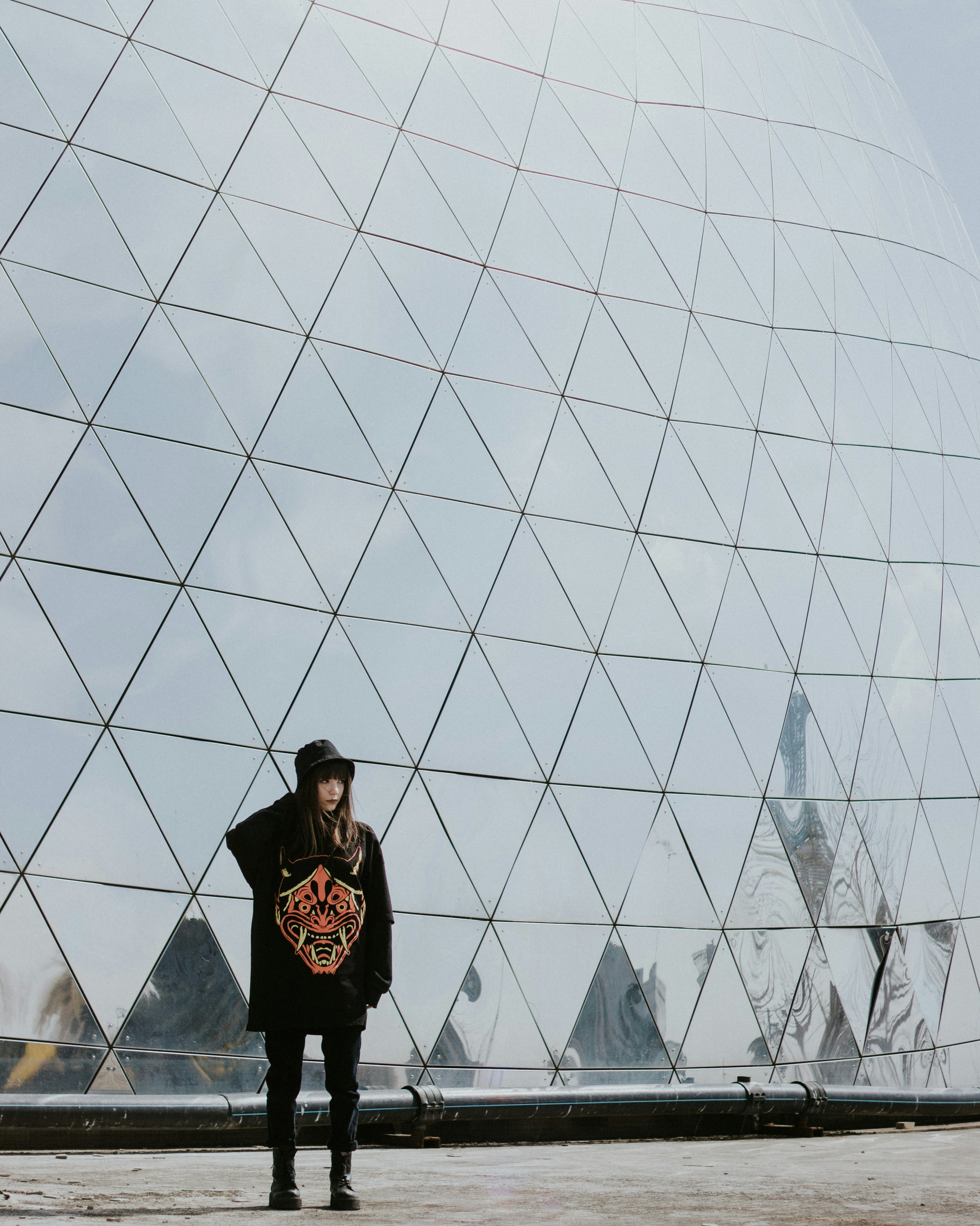 A figure stands confidently in front of a geometric glass structure, showcasing a bold graphic design on their coat. The reflective surface captures the surrounding environment.