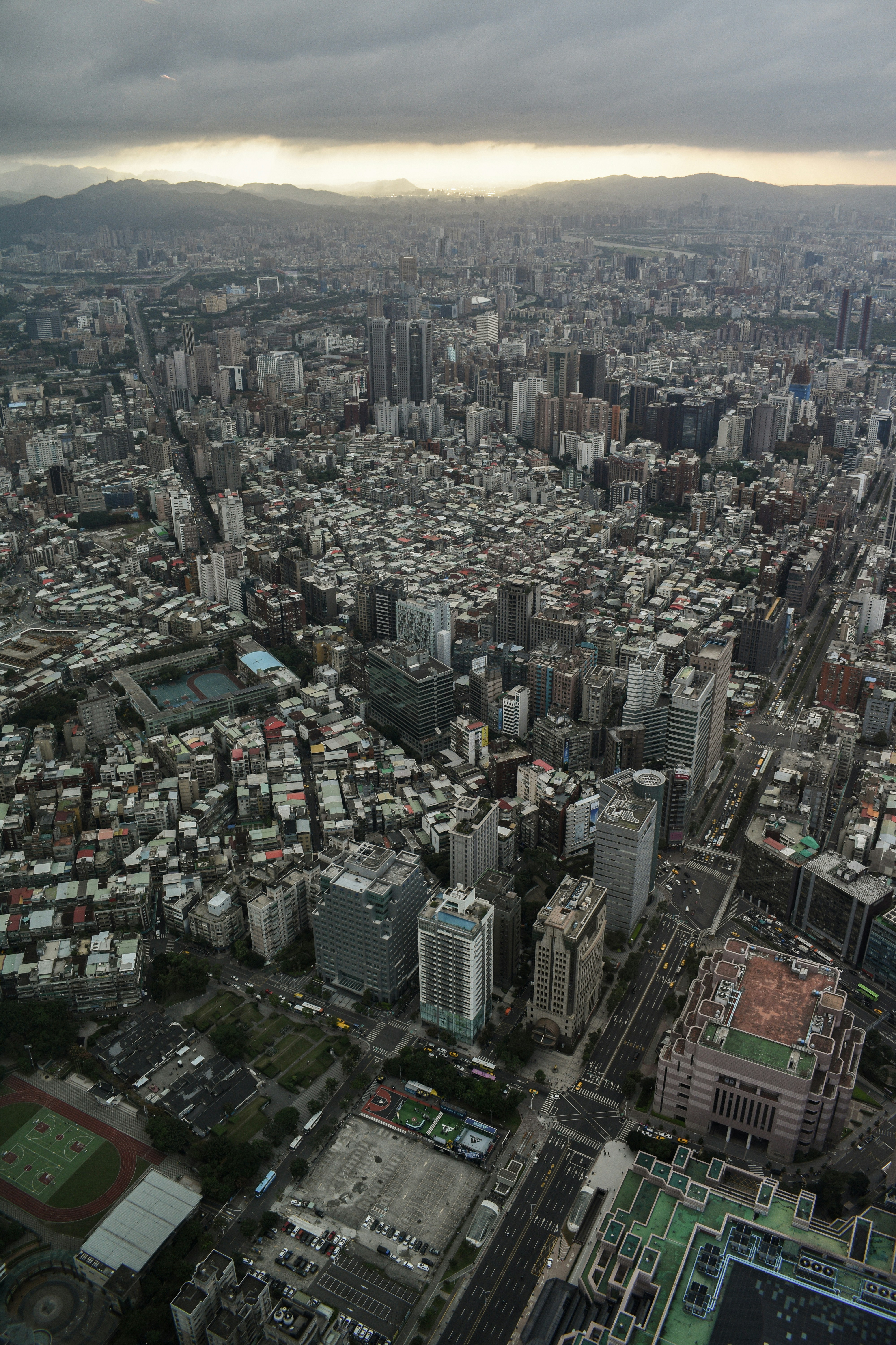 Aerial view of a sprawling urban landscape, showcasing a dense arrangement of buildings and streets, with mountains in the background. The scene captures the complexity of city life beneath a dramatic sky.
