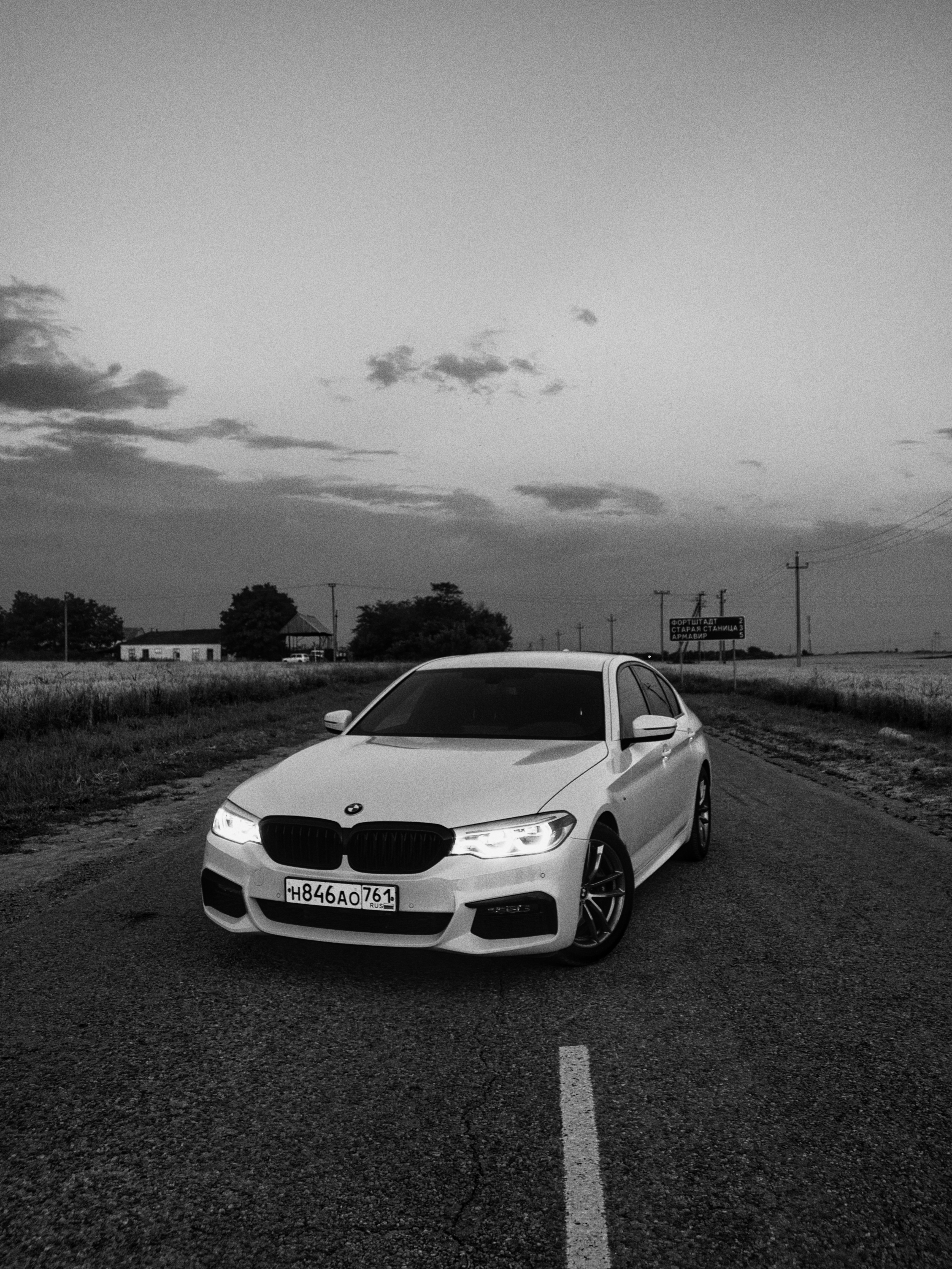 A sleek white BMW parked on a deserted road under a moody sky, highlighting its design and presence. The monochrome effect enhances the dramatic ambiance.