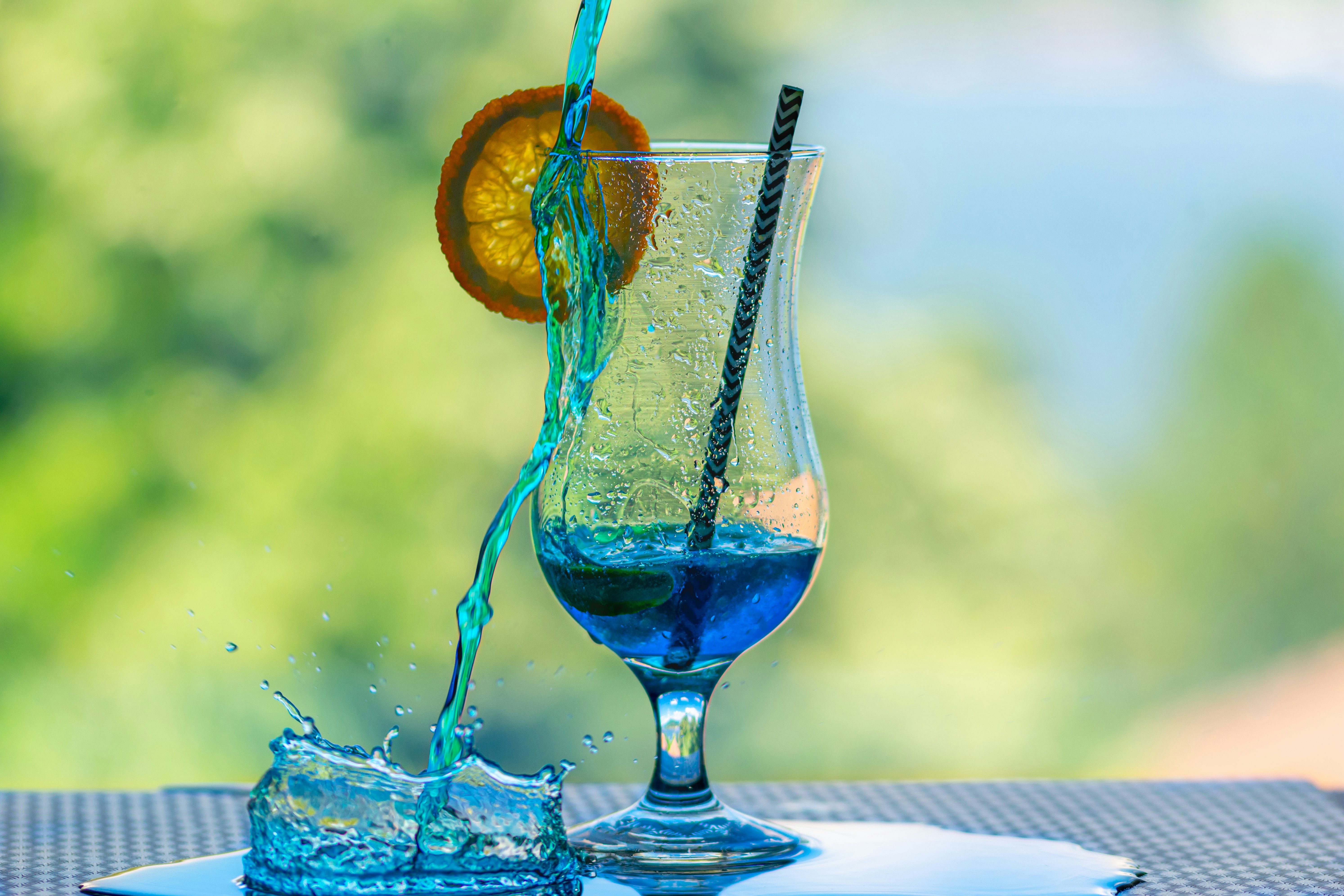 Vibrant blue cocktail with a splash cascading from the glass, garnished with an orange slice, set against a blurred green background.
