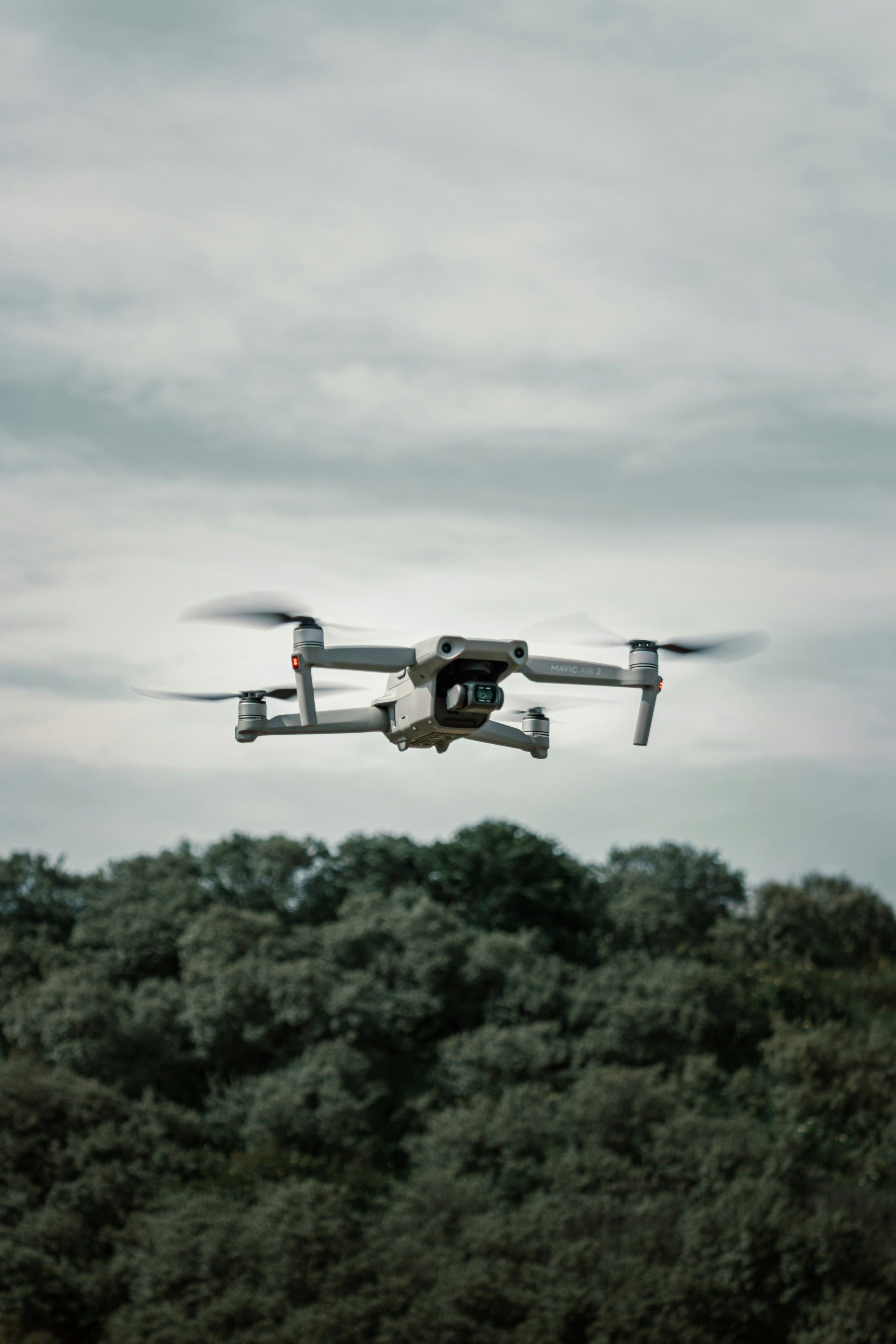 A drone hovers in the sky, capturing aerial views above a lush green forest under an overcast sky.