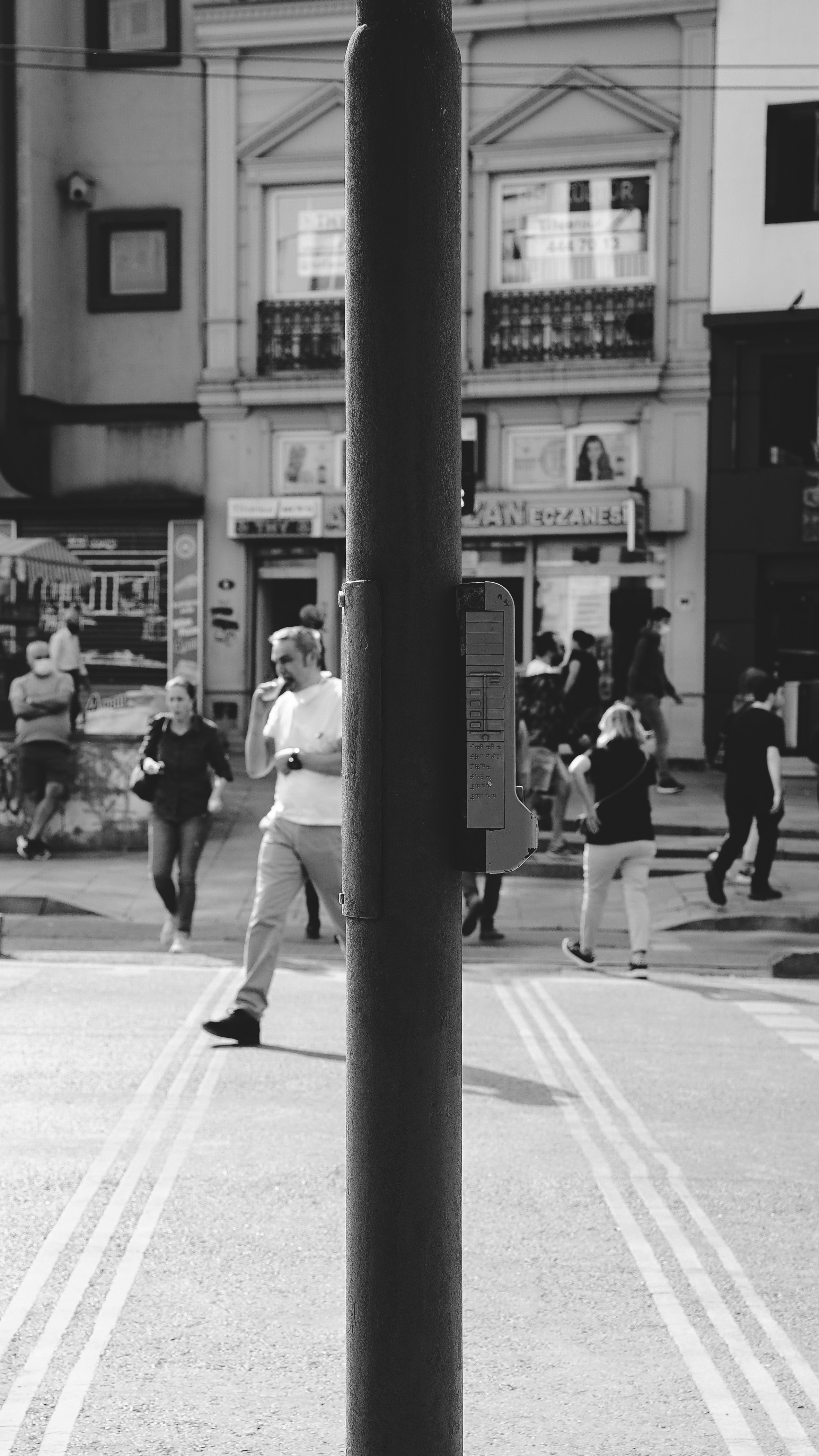 Busy street scene featuring pedestrians navigating a crosswalk, framed by a street pole. The background showcases urban architecture and vibrant storefronts.