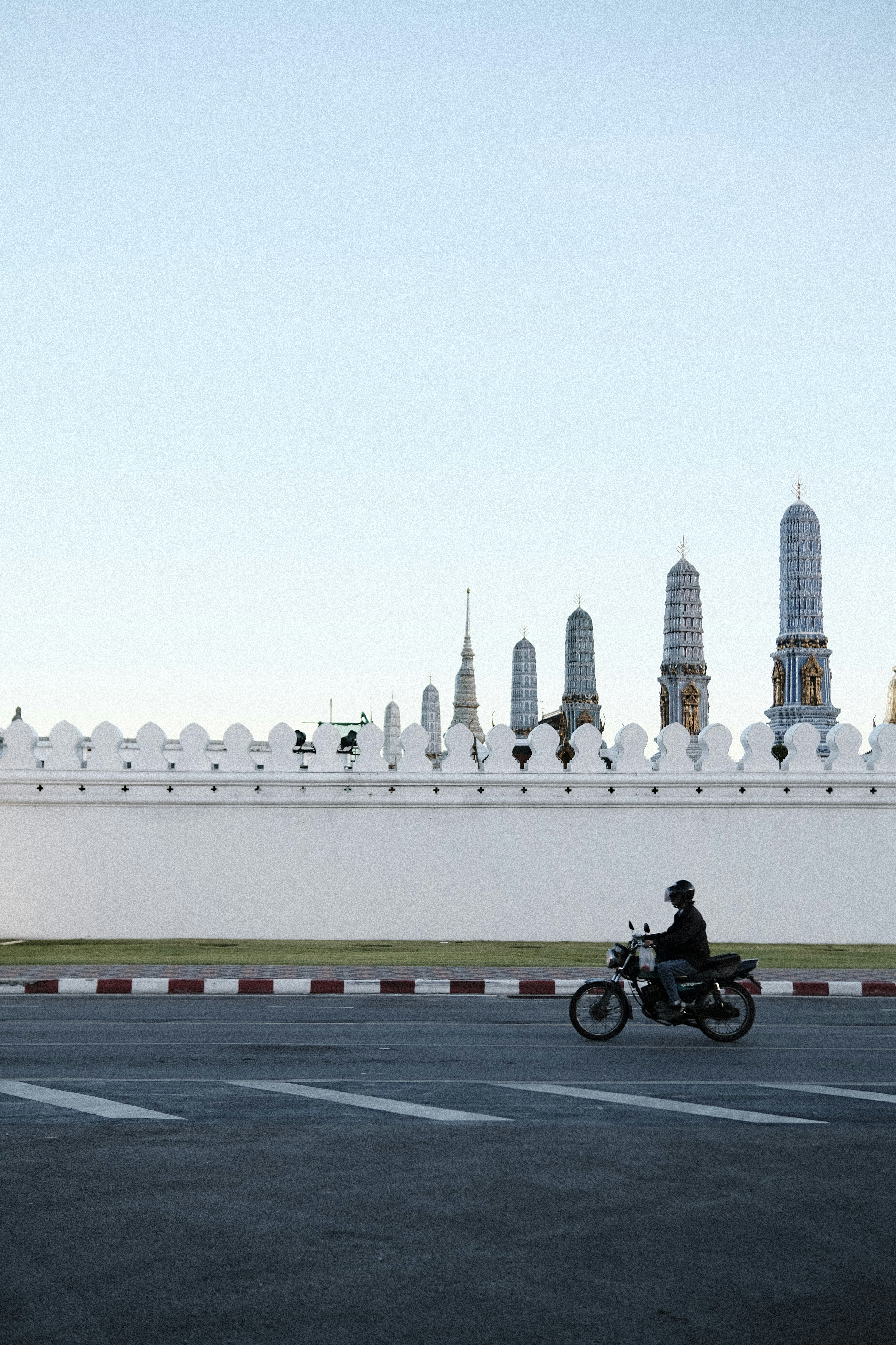 Motorcyclist navigating past a white wall adorned with intricate spires under a clear blue sky.