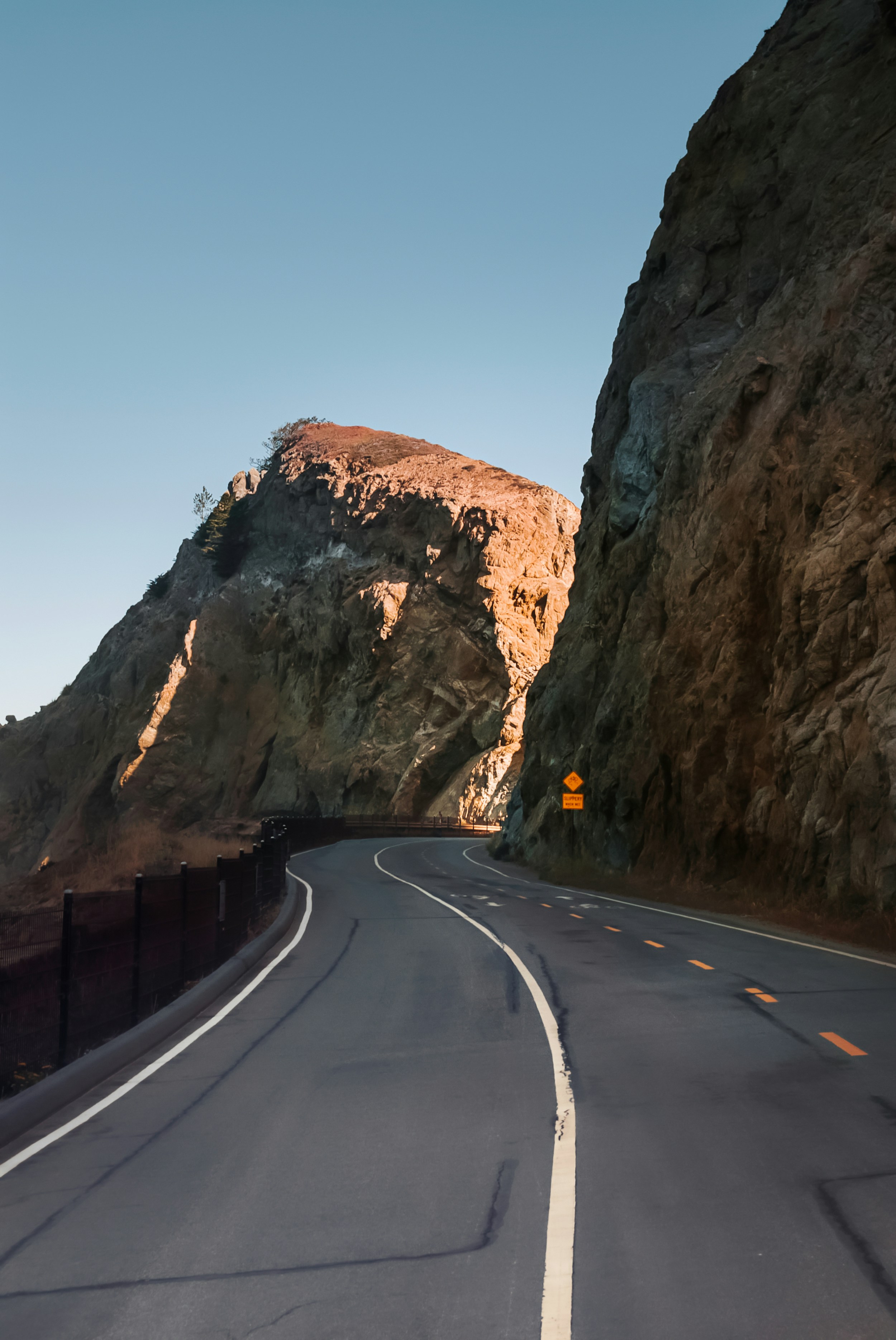 Curving road winding between steep, sunlit cliffs under a clear blue sky.
