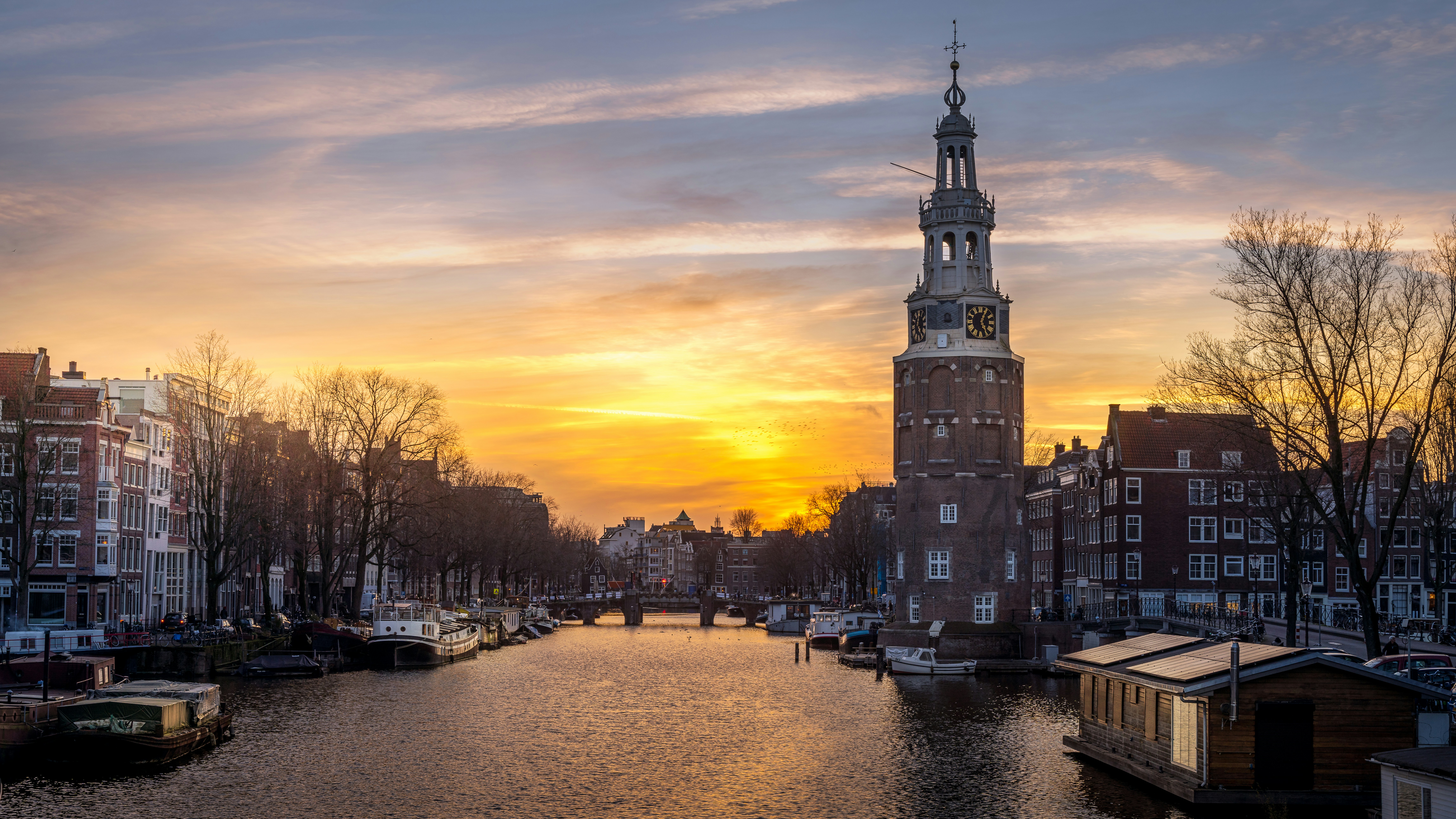 Historic clock tower overlooking a serene canal at sunset, with reflections shimmering on the water's surface.