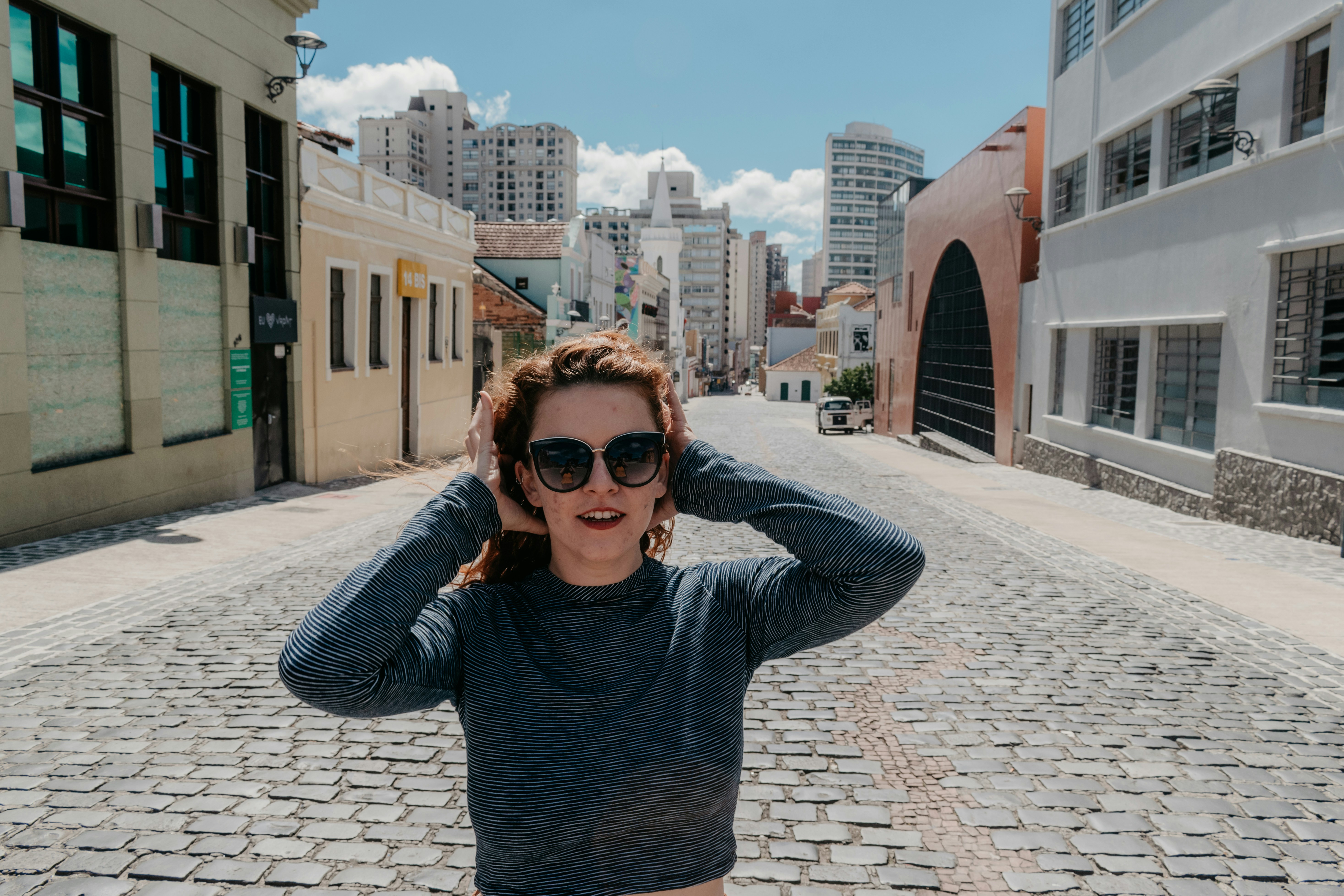 Woman with sunglasses stands on a sunny cobblestone street flanked by modern buildings.