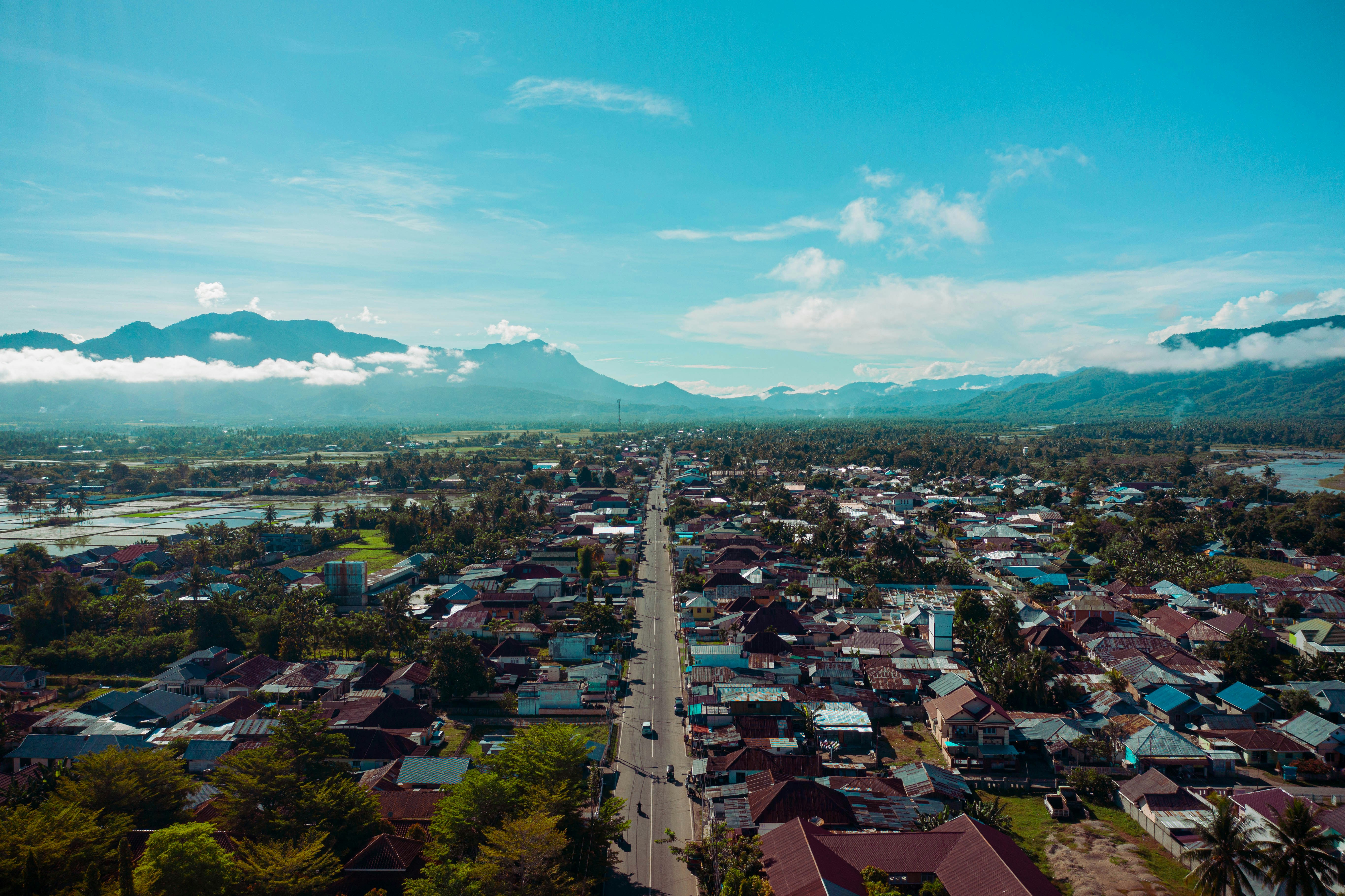 Aerial view of a bustling town bordered by lush mountains and clear skies, showcasing the vibrant rooftops and a central road leading through the community.