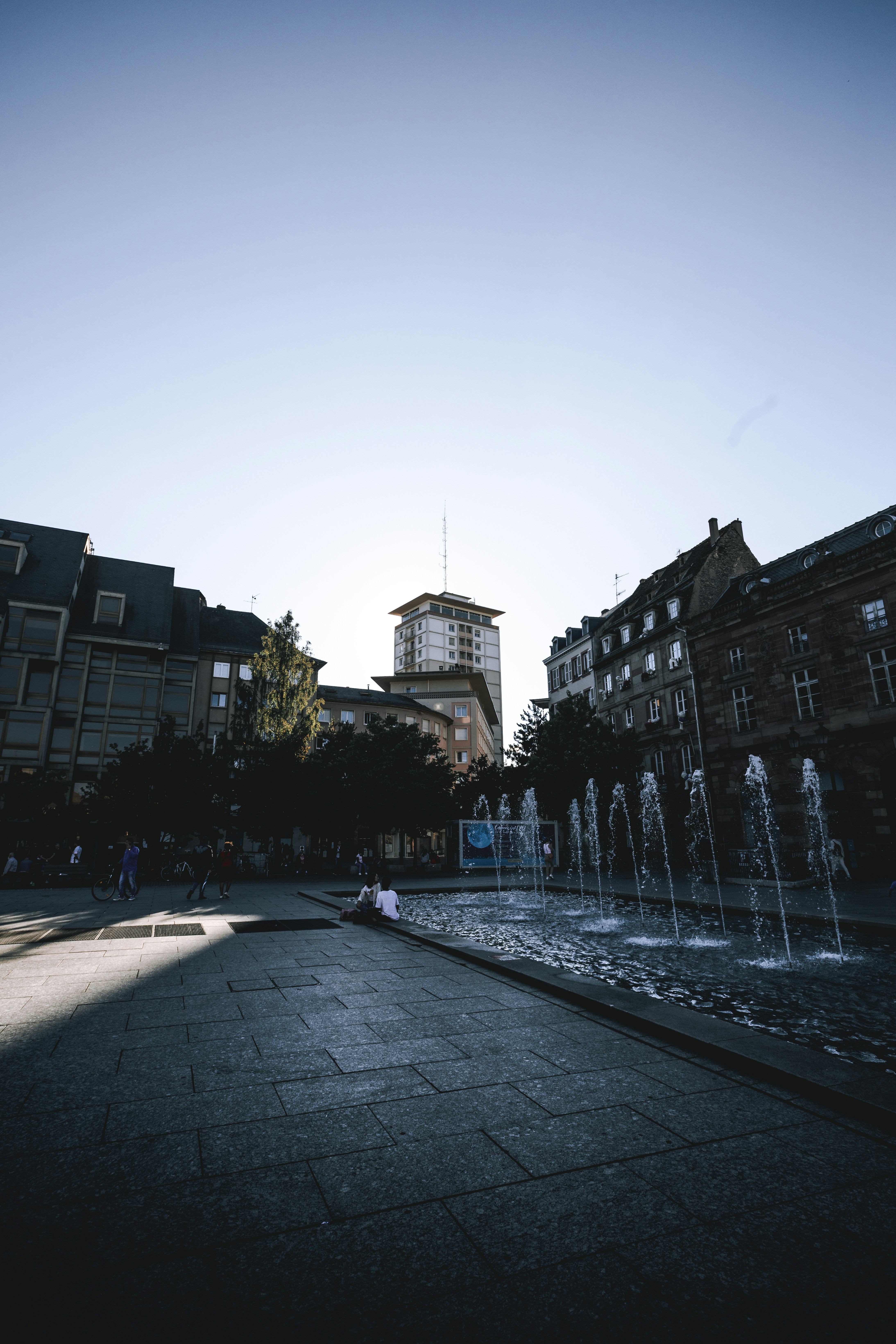 Fountain splashing in a vibrant city square surrounded by modern and historic architecture. People relax and enjoy the ambiance.