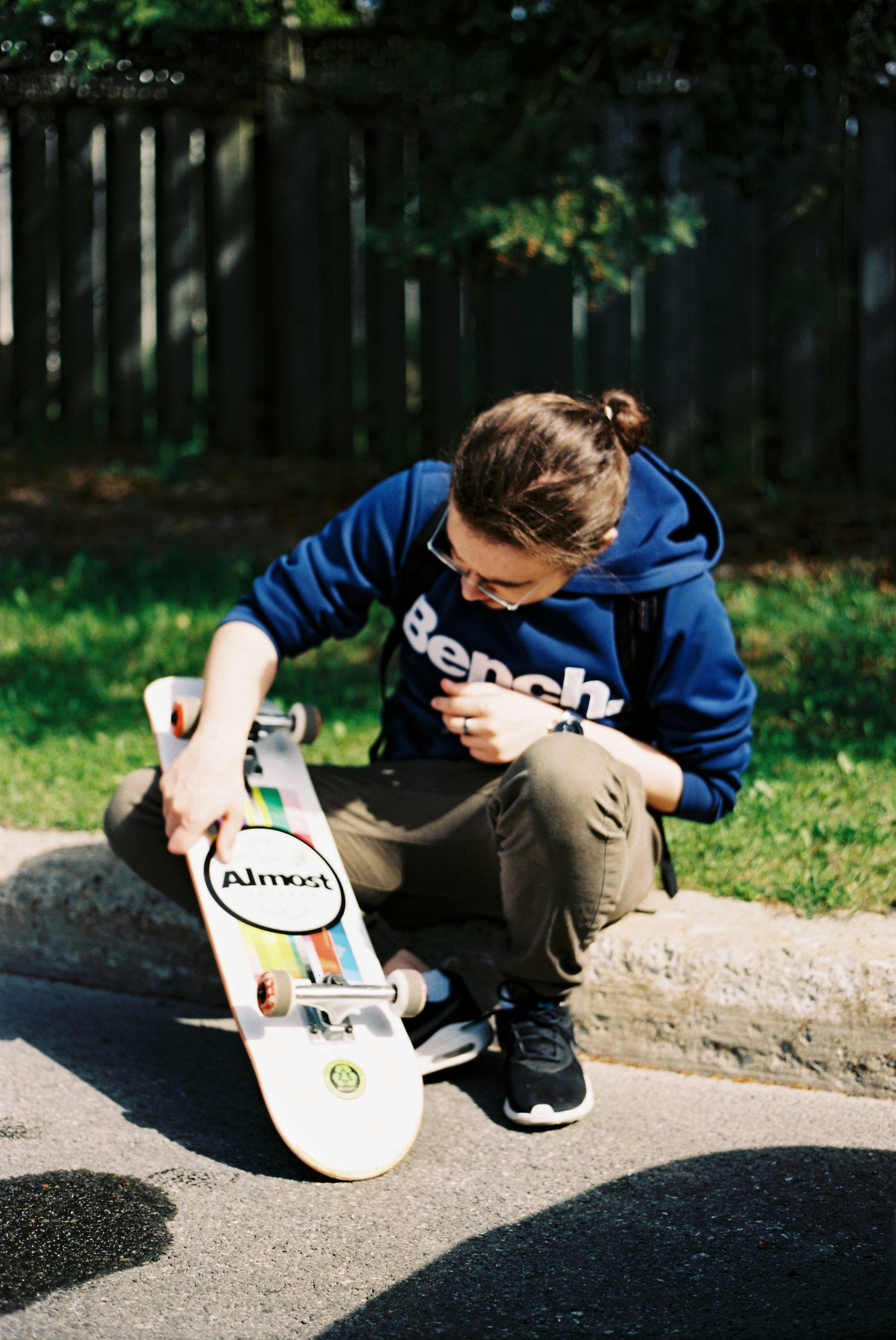 Sunlit Pause: A Skater in a Blue Hoodie Adjusts an Almost Deck on the Curb