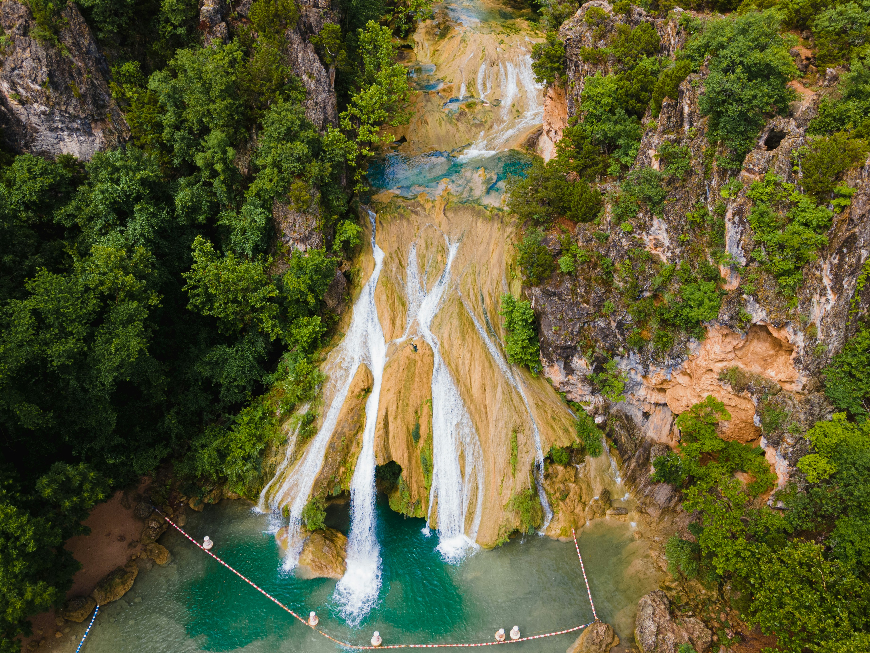 Aerial view of a vibrant waterfall cascading over rocky terrain, surrounded by lush greenery and a tranquil turquoise pool below.