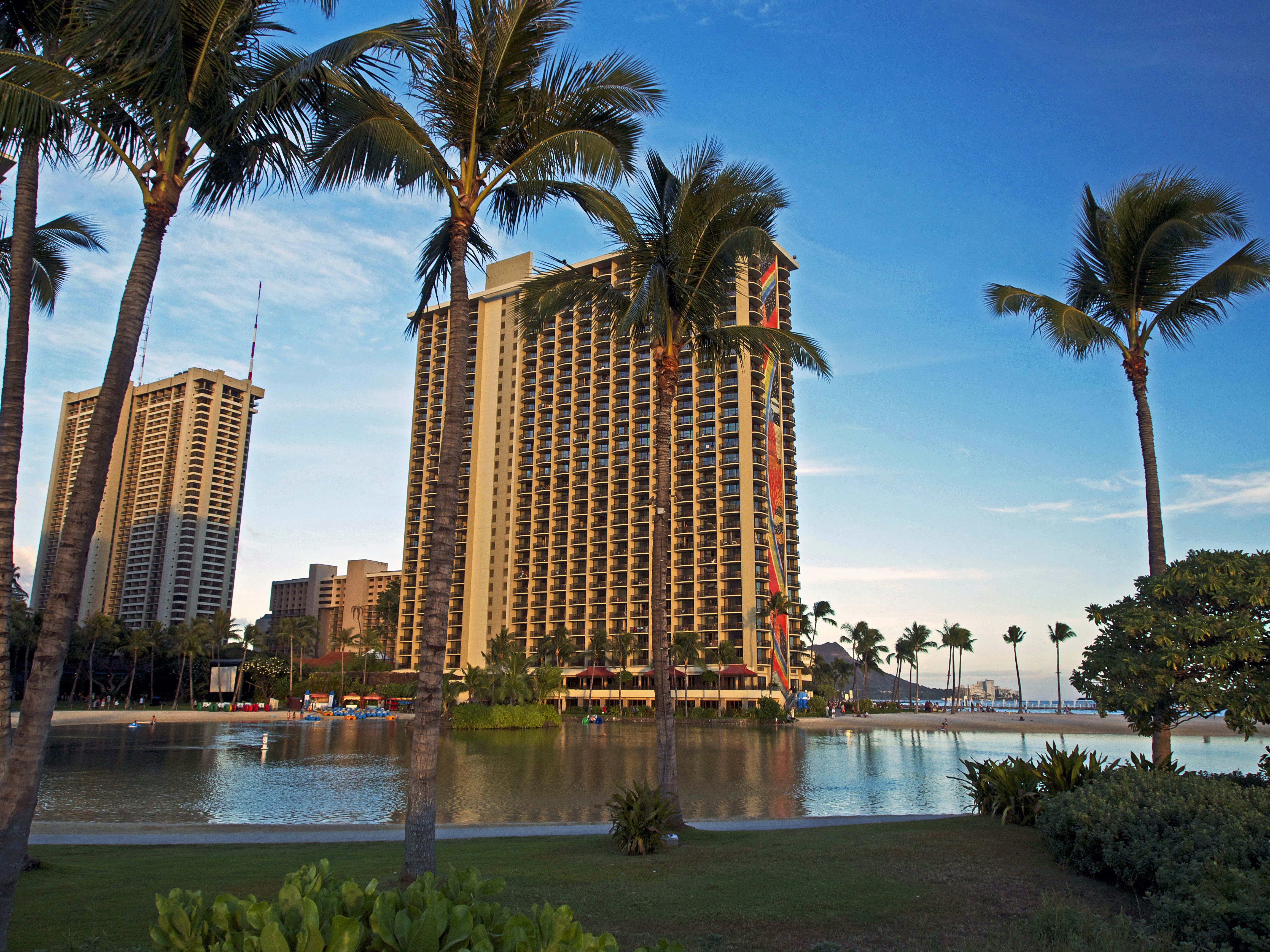 Tall palm trees frame a vibrant cityscape with a shimmering lake in the foreground during sunset.