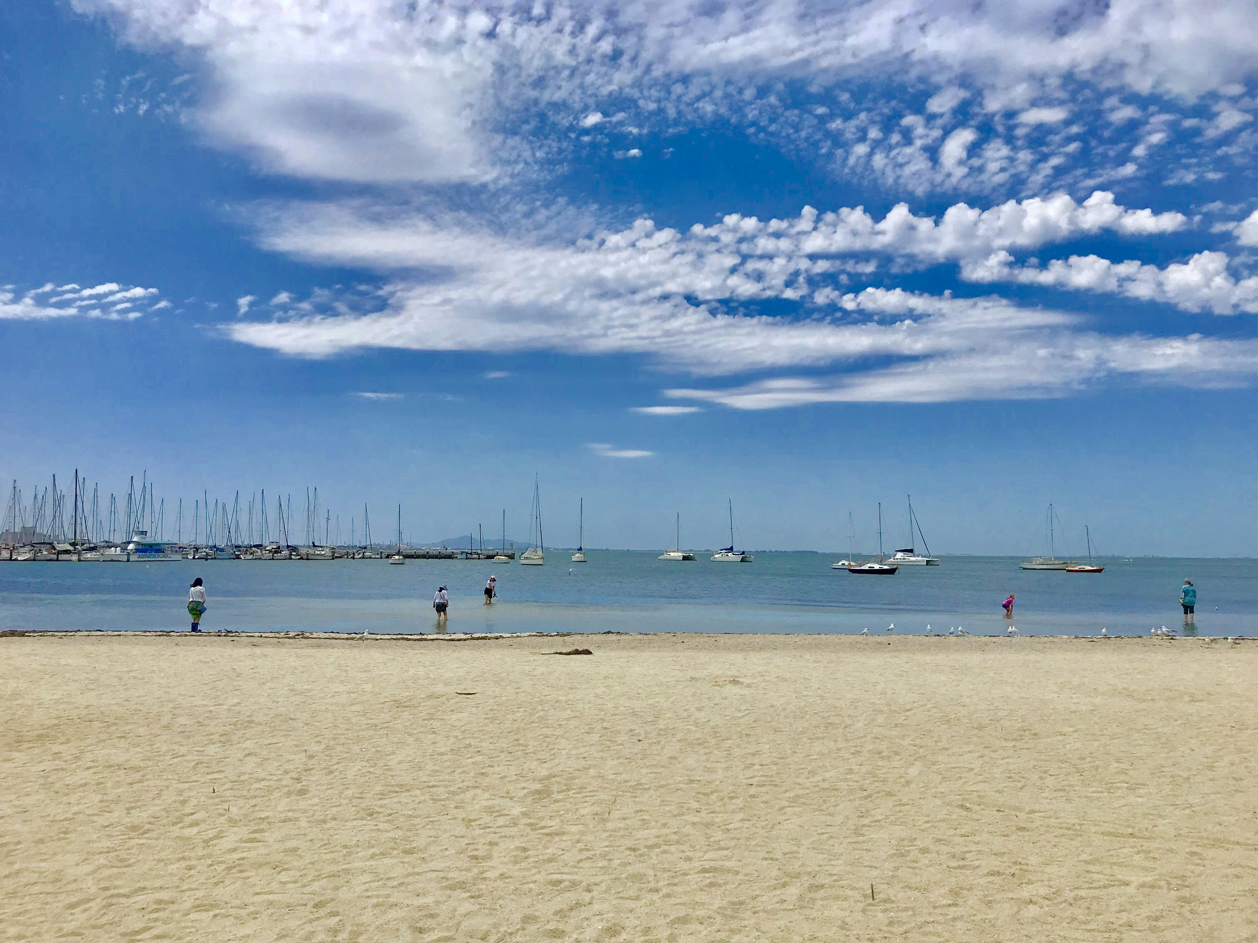 Bright sandy beach with scattered beachgoers and sailboats against a blue sky with wispy clouds.